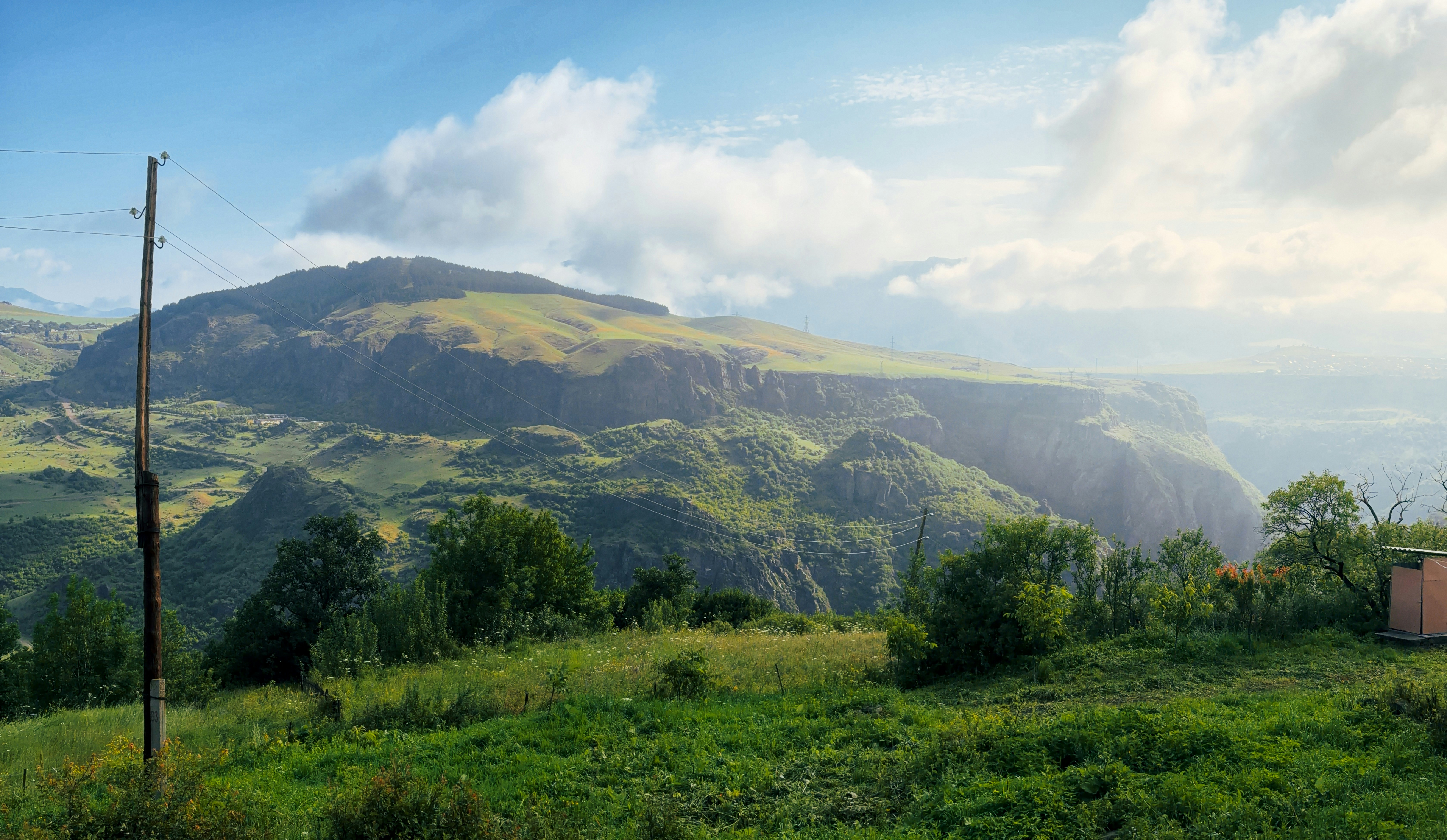 Rolling green hills and distant cliffs under a bright, slightly hazy sky; a quiet landscape with a lone utility pole and scattered shrubs add depth.