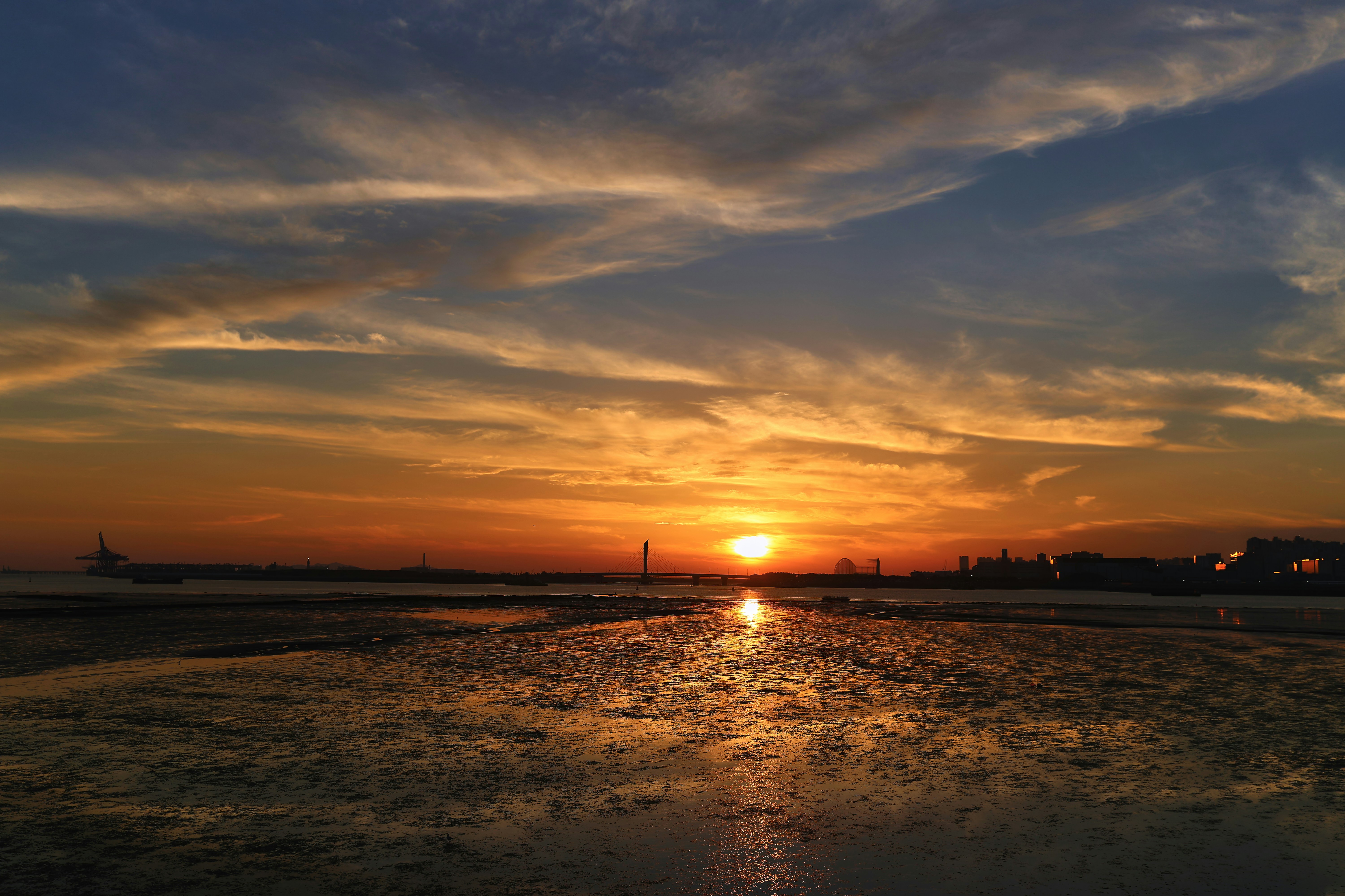 Sunset over a calm coastal water with a glowing horizon and silhouetted skyline; warm reflections on the shallow tide. A serene seascape capturing the sun dipping toward the city.