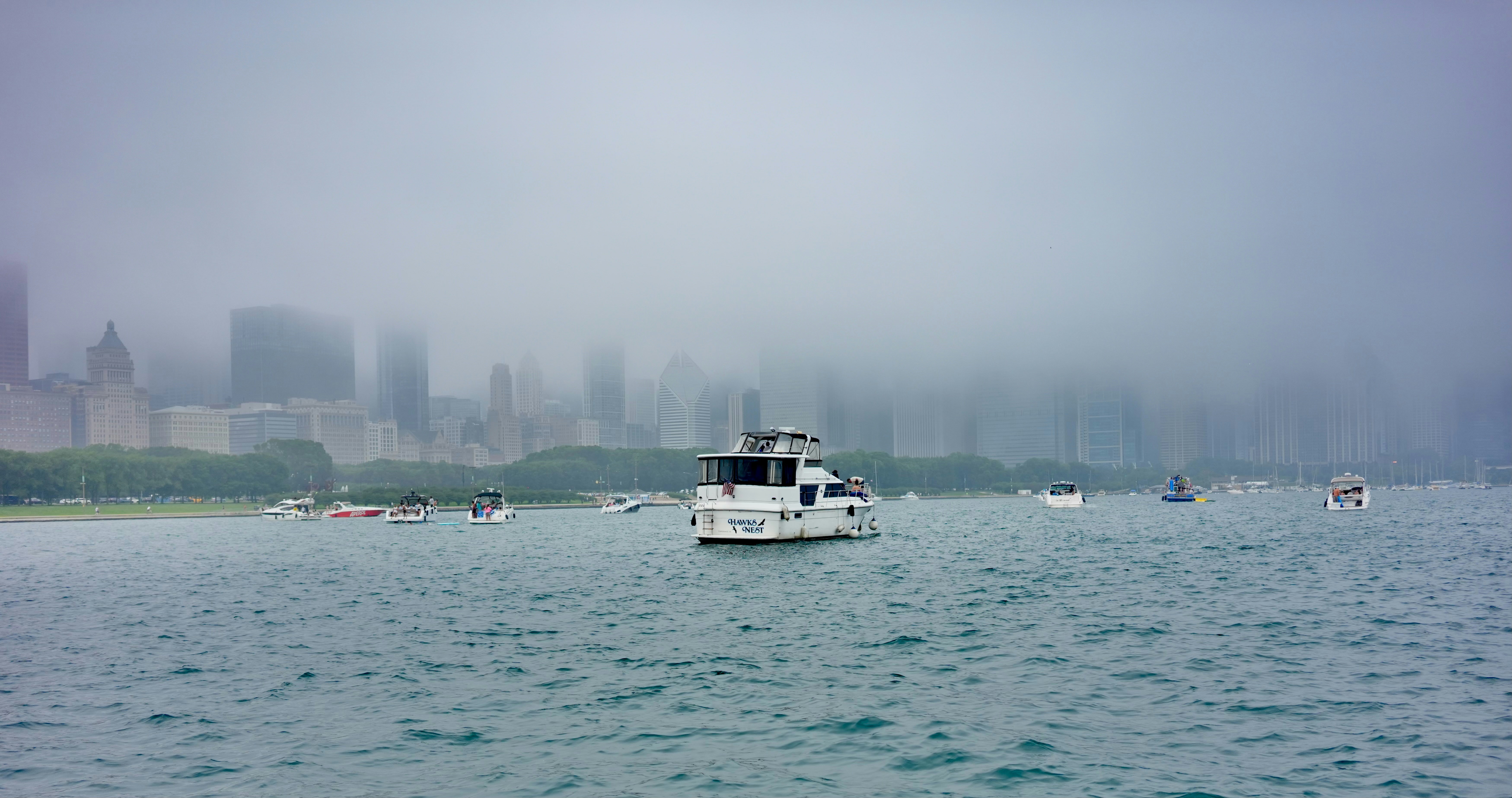 A white motorboat anchors in a calm harbor with a distant urban skyline veiled by fog, creating a serene maritime scene.