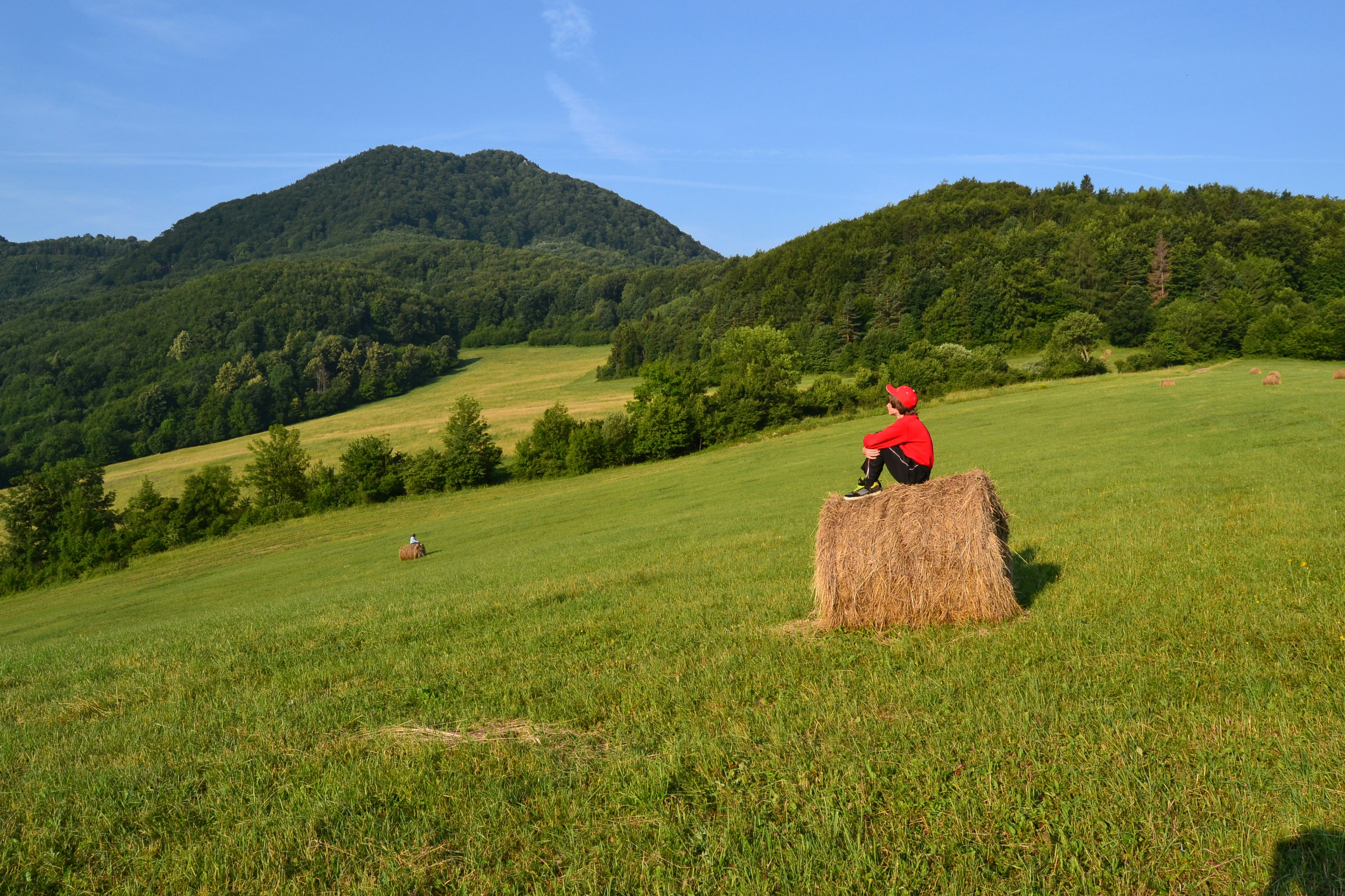 Red-Capped Figure on a Hay Bale Amid Rolling Hillscape