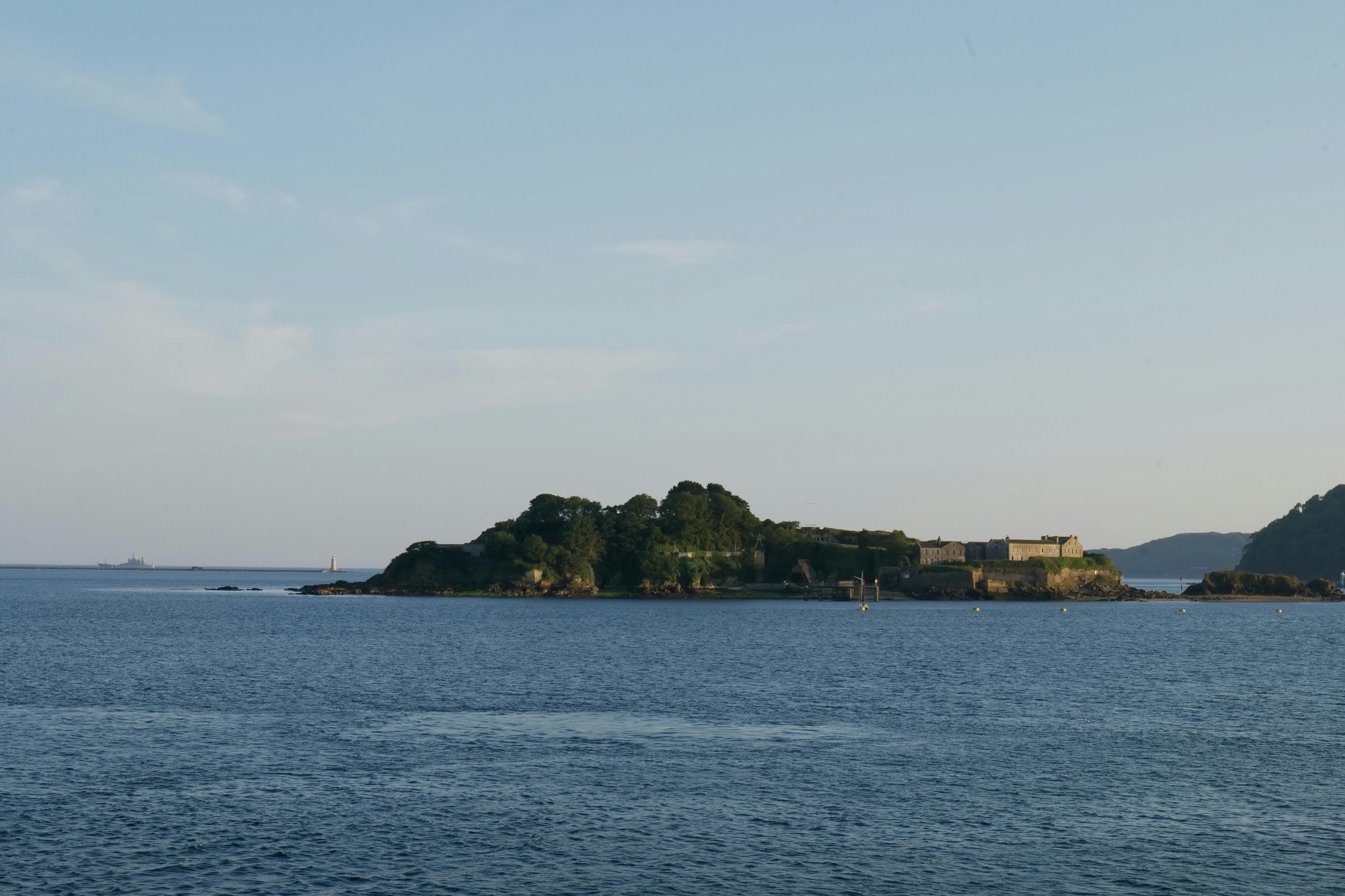 A calm seascape shows a small tree-covered island with a historic fortification along the shore, resting on placid blue water under a clear sky.