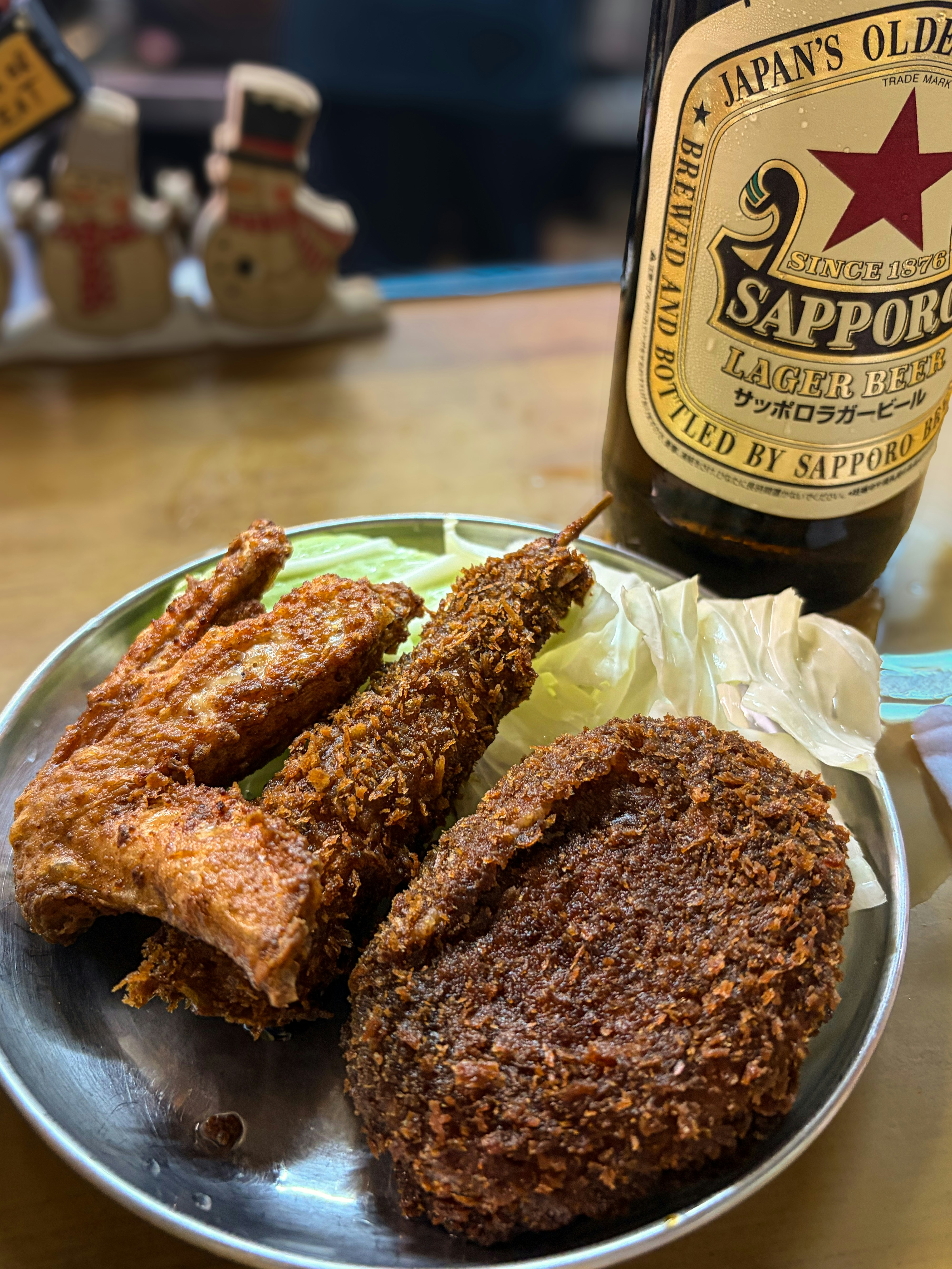 Crispy fried chicken and croquettes rest on a metal plate beside a bottle of Sapporo beer; lettuce leaves provide contrast as a casual dining setup.