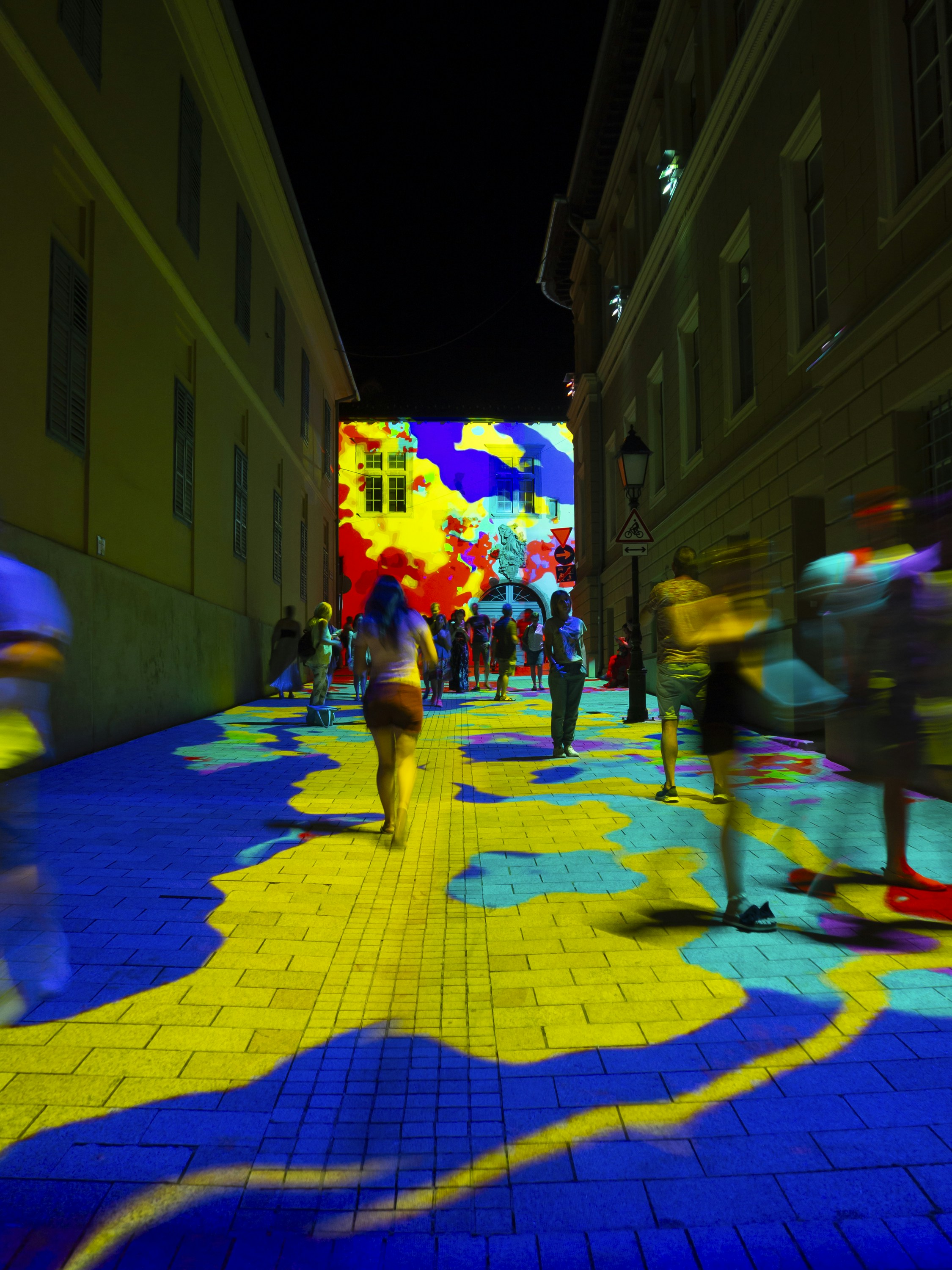 Vibrant light projection floods a narrow street with abstract colors as silhouettes of pedestrians move through the scene.