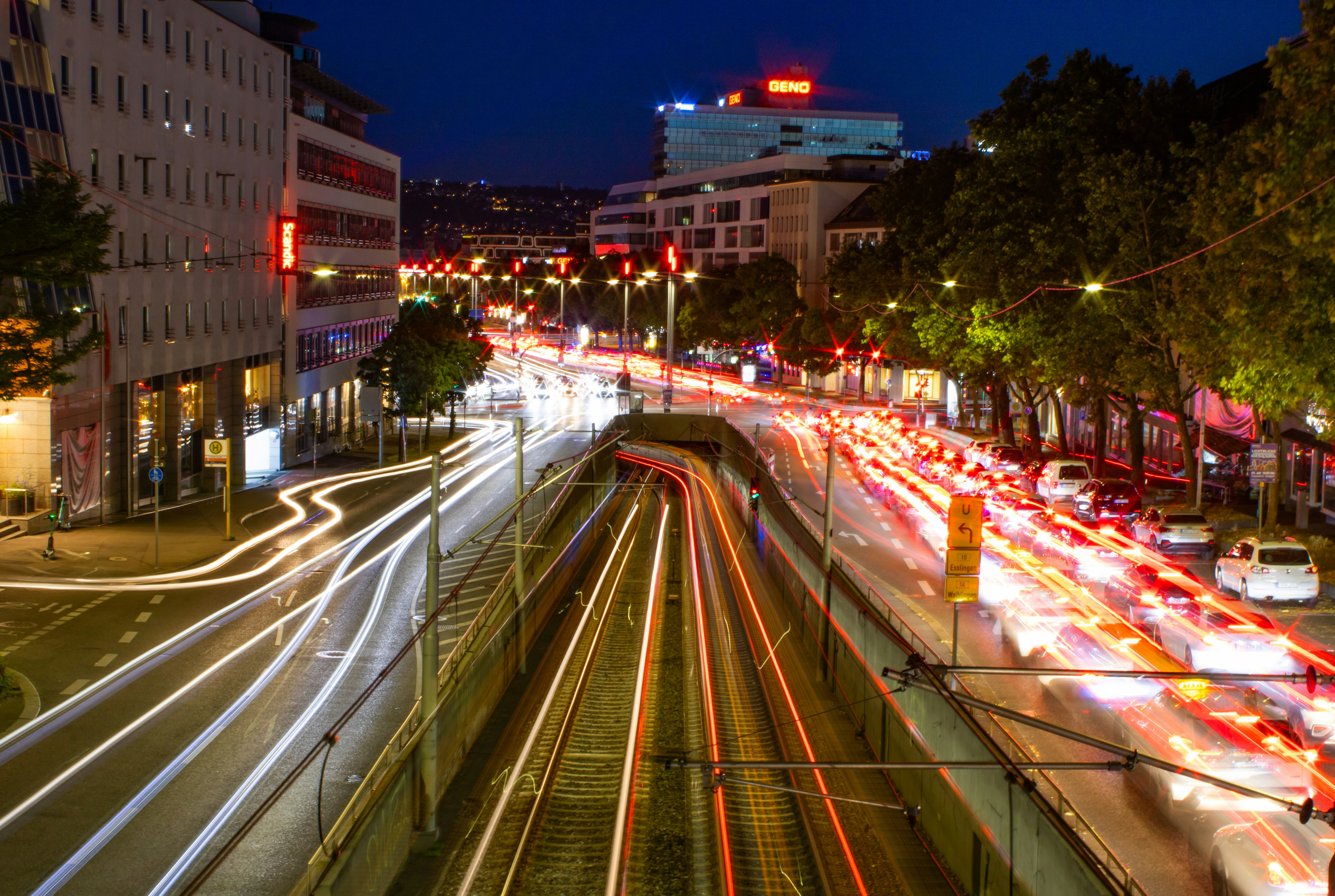 Long-exposure view of a city street and rail lines at night, capturing light trails from moving vehicles along a central spine of tracks with surrounding buildings and trees. The image emphasizes motion and urban glow.