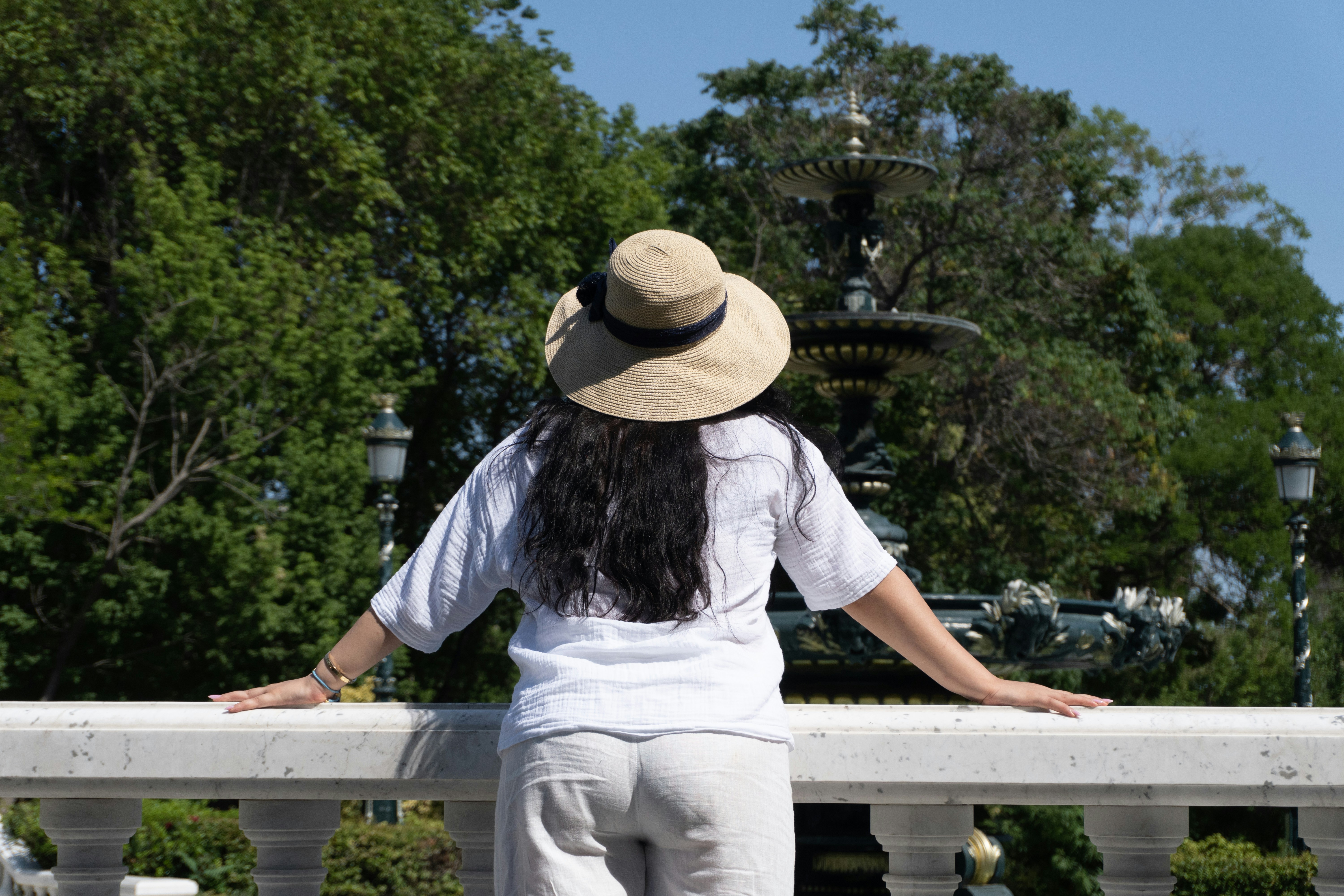 Woman in a white outfit with a wide-brim hat leaning on a fountain railing