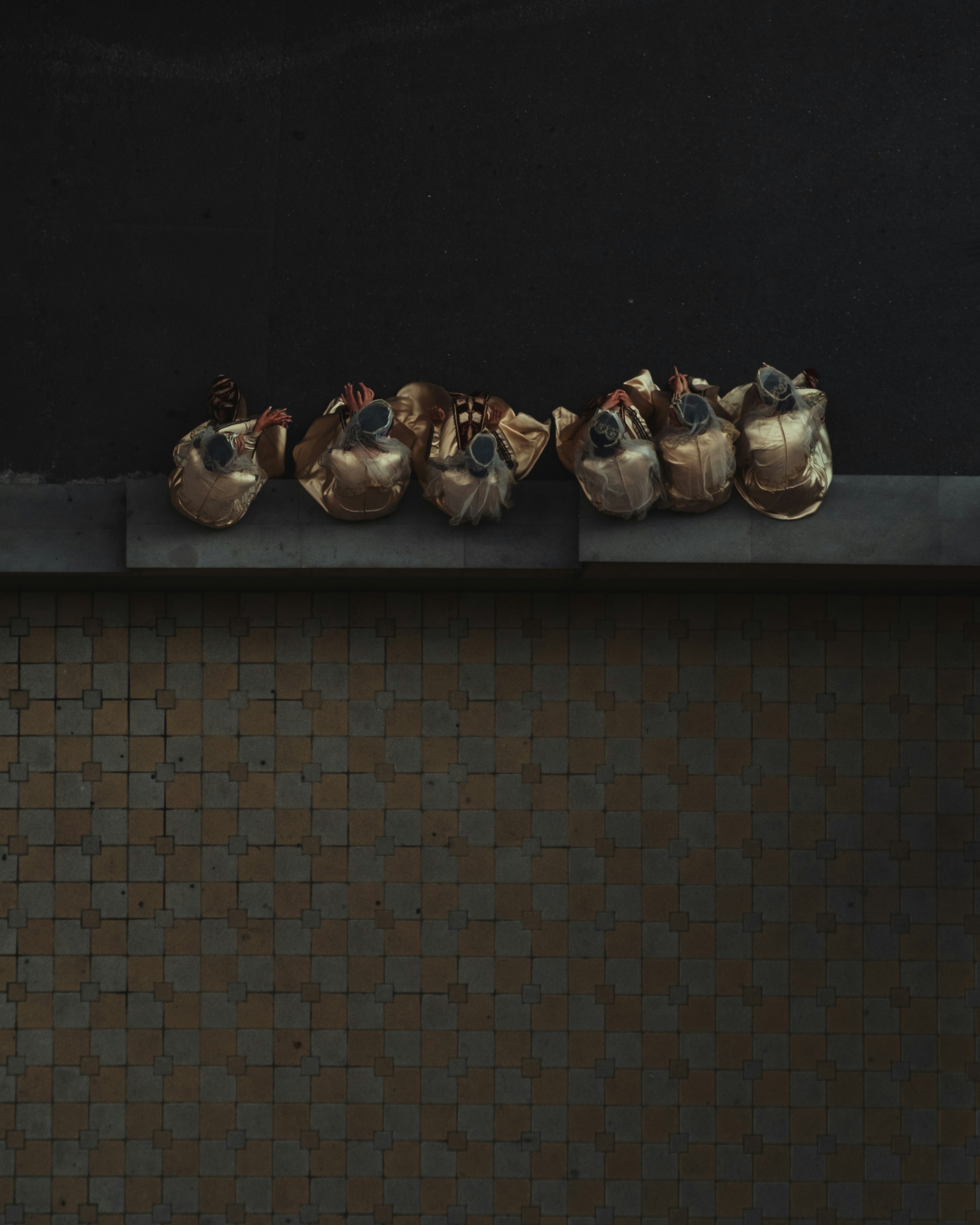 A row of gold-toned figures rests along a concrete edge above a tiled wall, their backs turned to the viewer in a dimly lit scene. This is a photographic capture, not a painting or illustration.