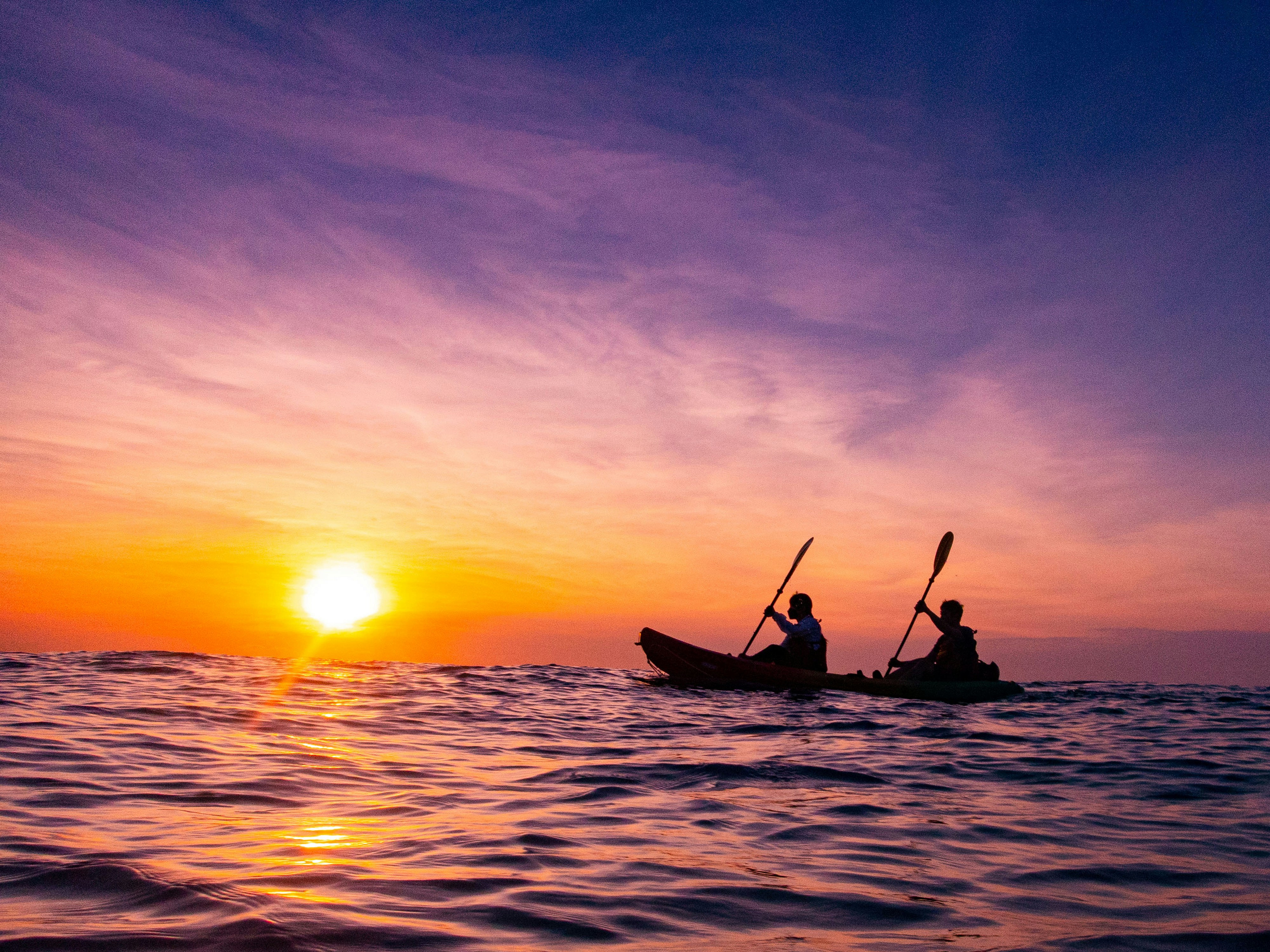 Two kayakers paddle across a calm sea at sunset, their silhouettes contrasting against the orange-pink sky and glowing horizon.