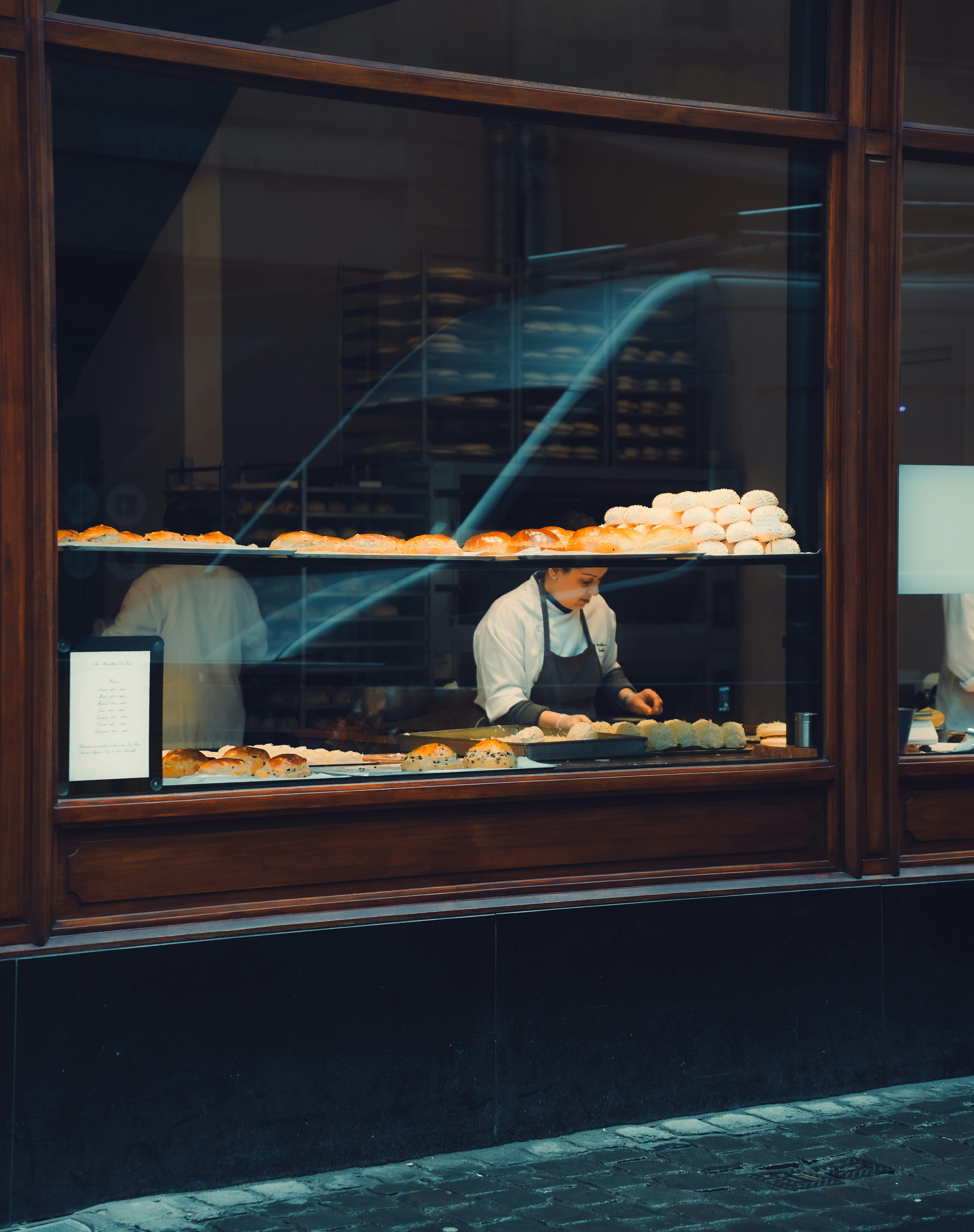 A bakery worker arranges pastries behind a shop window, with rows of bread and buns visible along a curved display; clean wooden frame and glass reflections frame the scene.