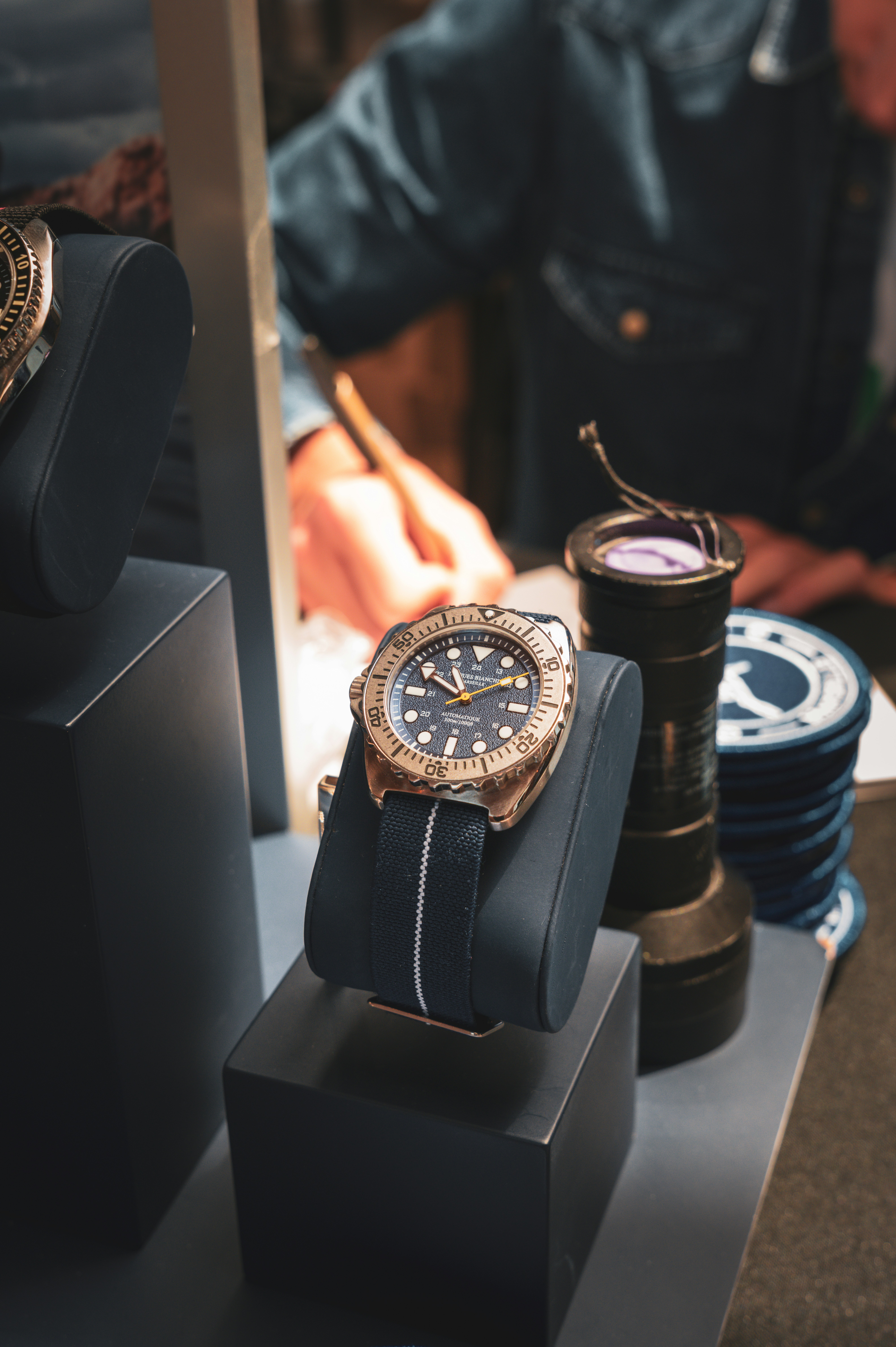 A close up of a dive watch displayed on a stand at a watch event, captured with shallow depth of field. Warm lighting and a blurred background highlight the texture of the dial, bronze case, and NATO strap.
