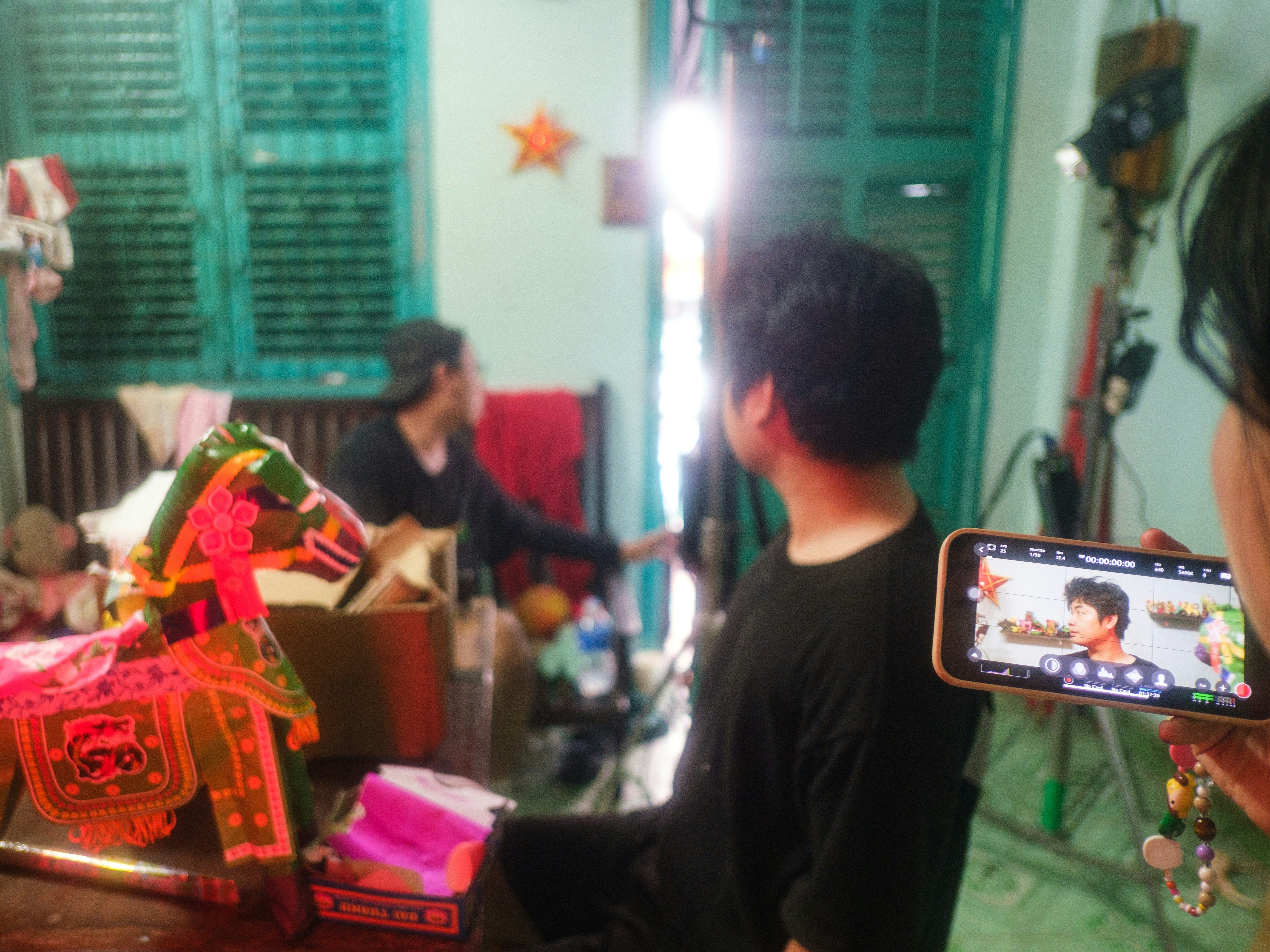Portrait in a cluttered studio: a young man faces away as a livestreaming phone captures the scene