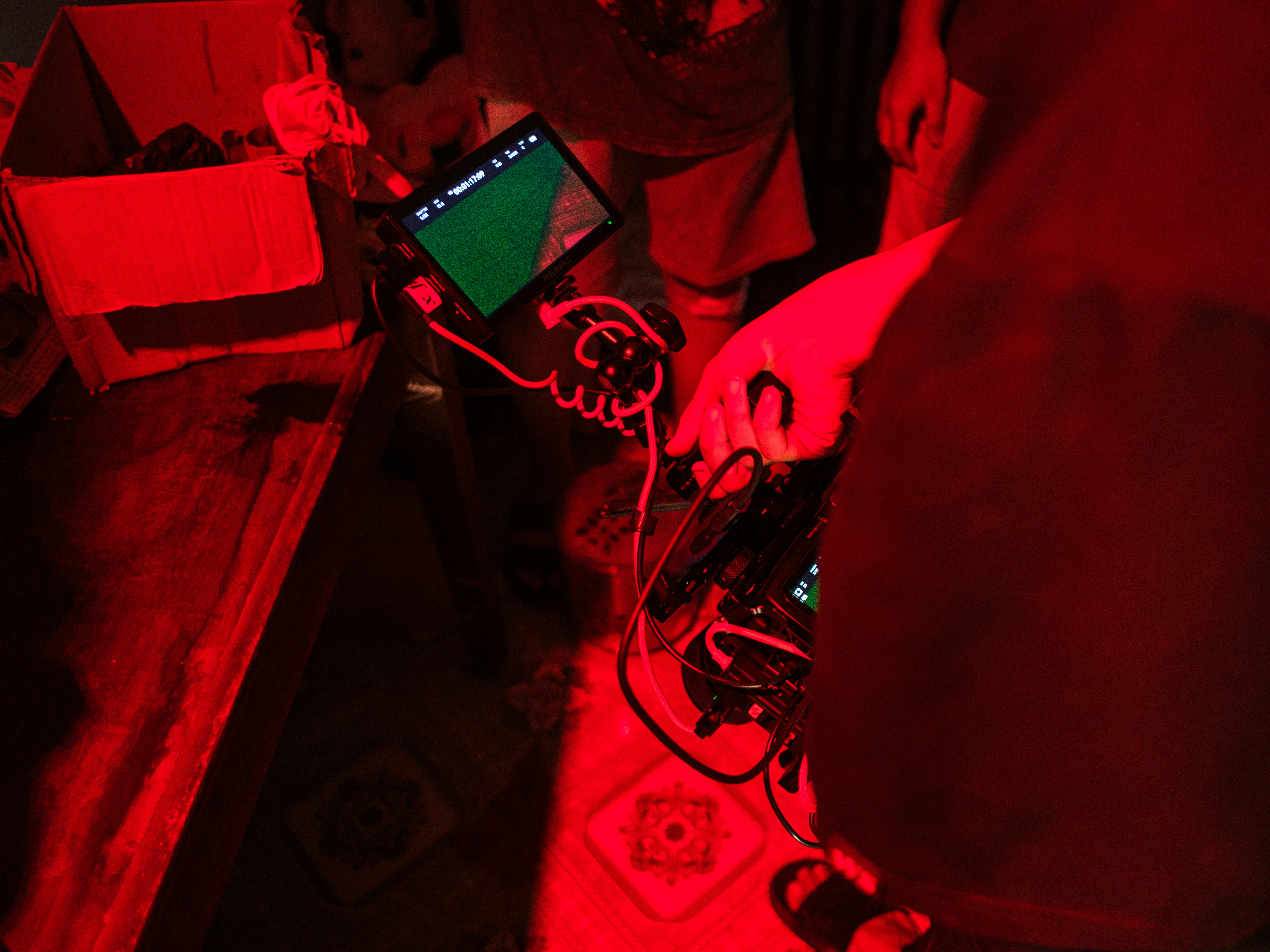 Close-up of hands manipulating electronic gear under red lighting, with a small screen and cables visible on a cluttered table.