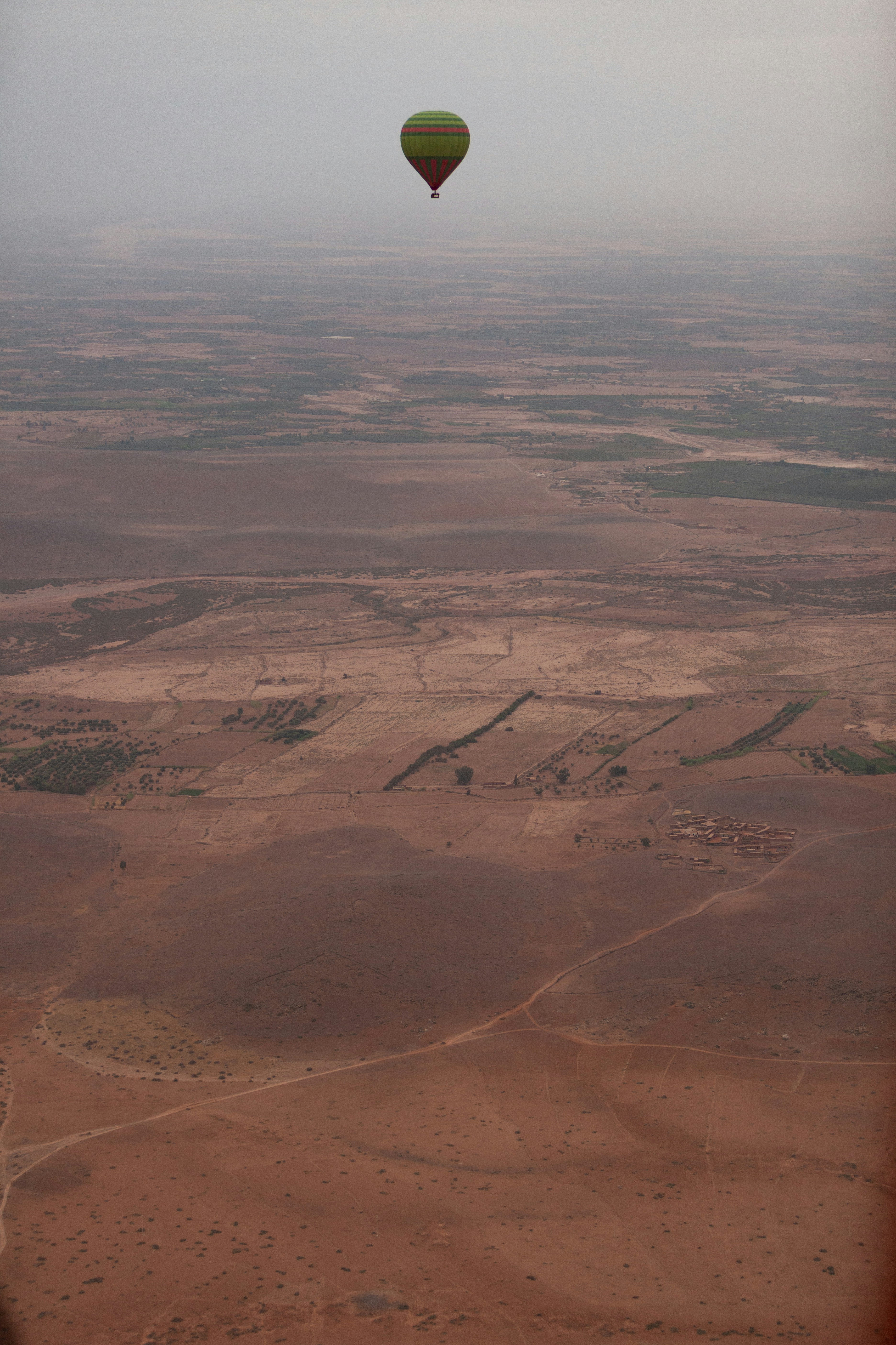 A lone hot air balloon drifts over a vast, arid landscape with patchwork fields and winding roads below; the scene emphasizes scale and solitude in a muted palette.