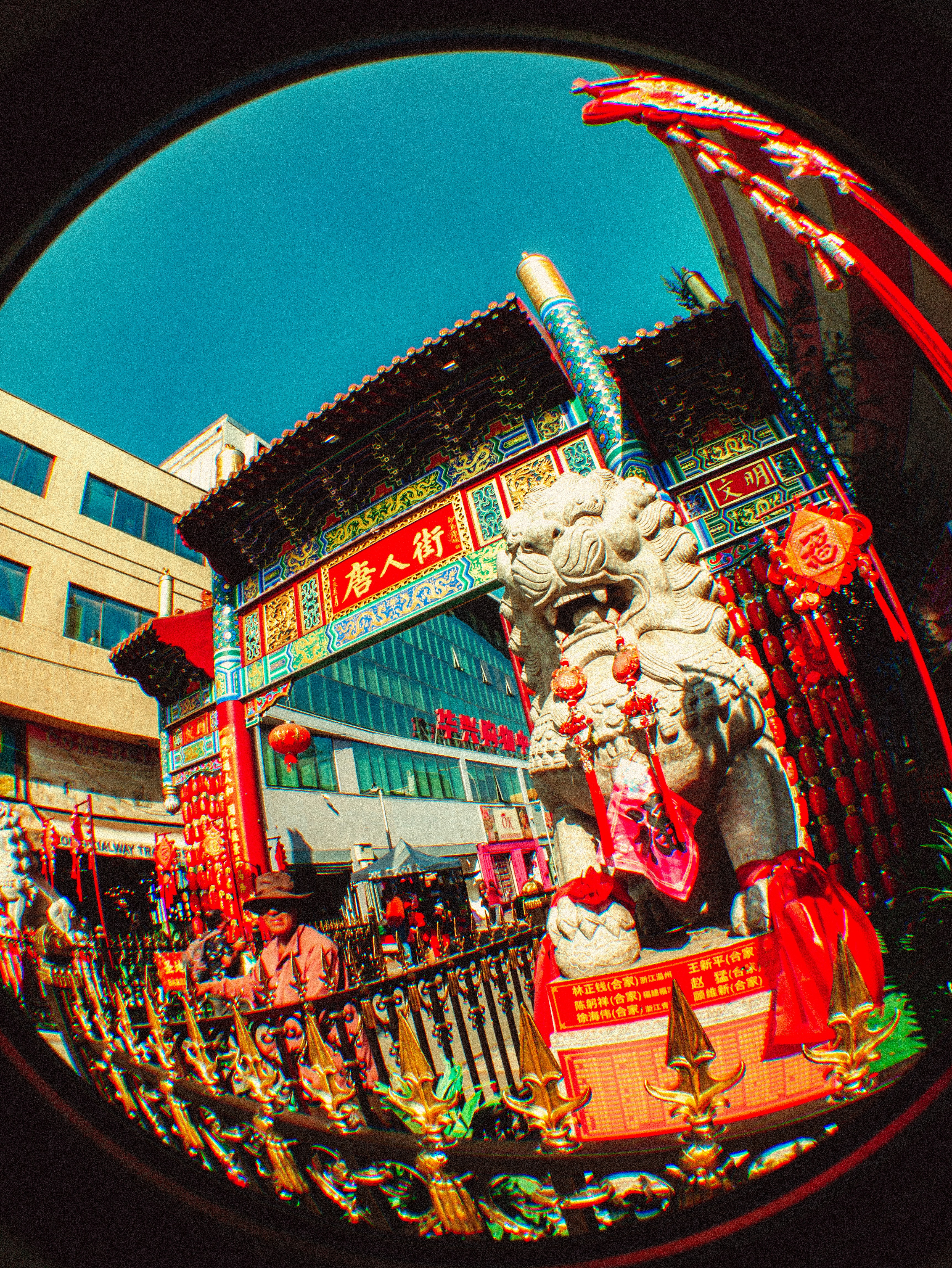 Vibrant Chinatown festival scene framed through a circular fisheye lens, highlighting the ornate archway, lion statue, and red lanterns against a bright urban backdrop.