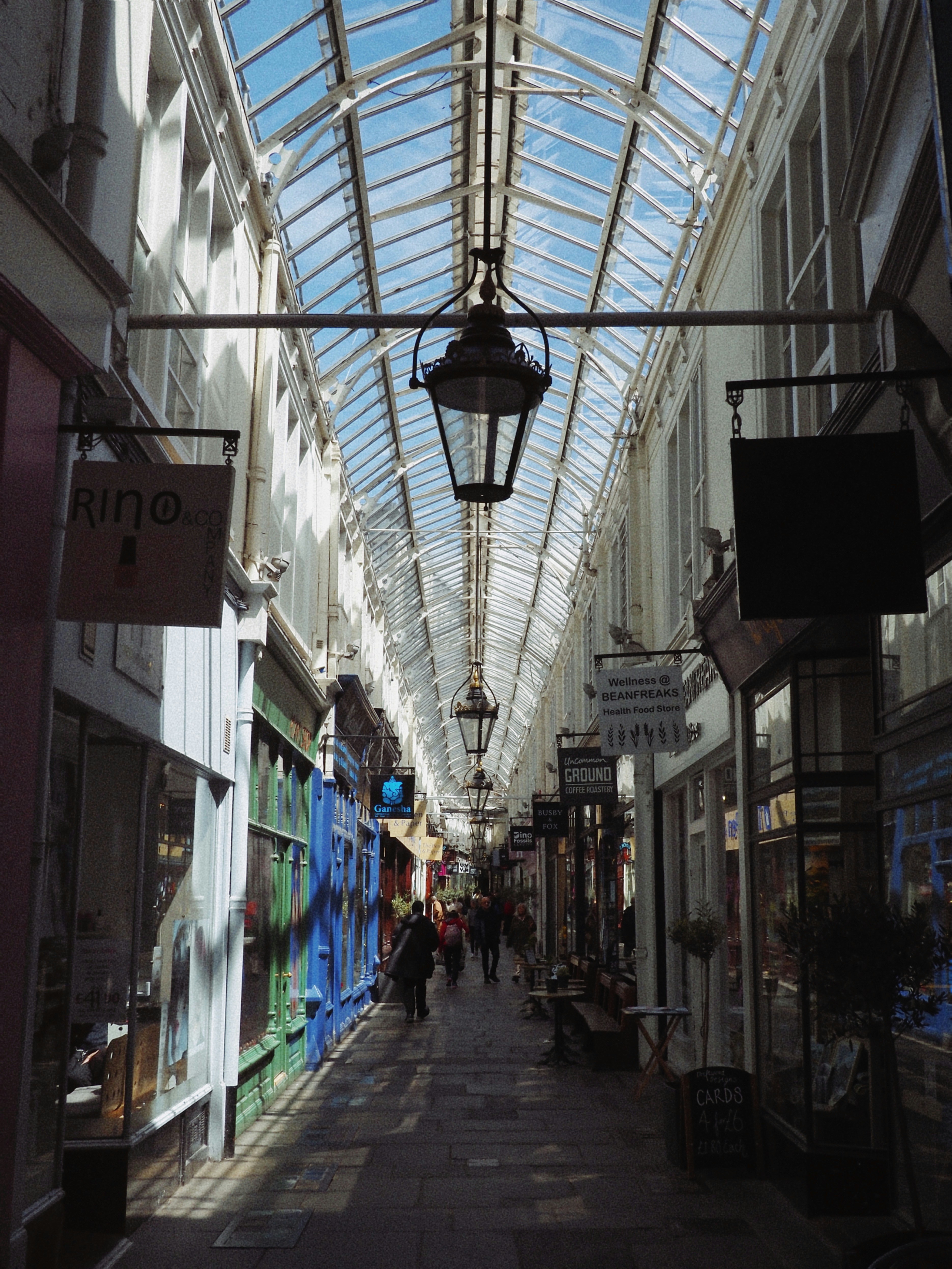 Cozy indoor shopping arcade with a glass-paneled roof, market storefronts lining a cobblestone walkway, and pedestrians beneath a central lantern.