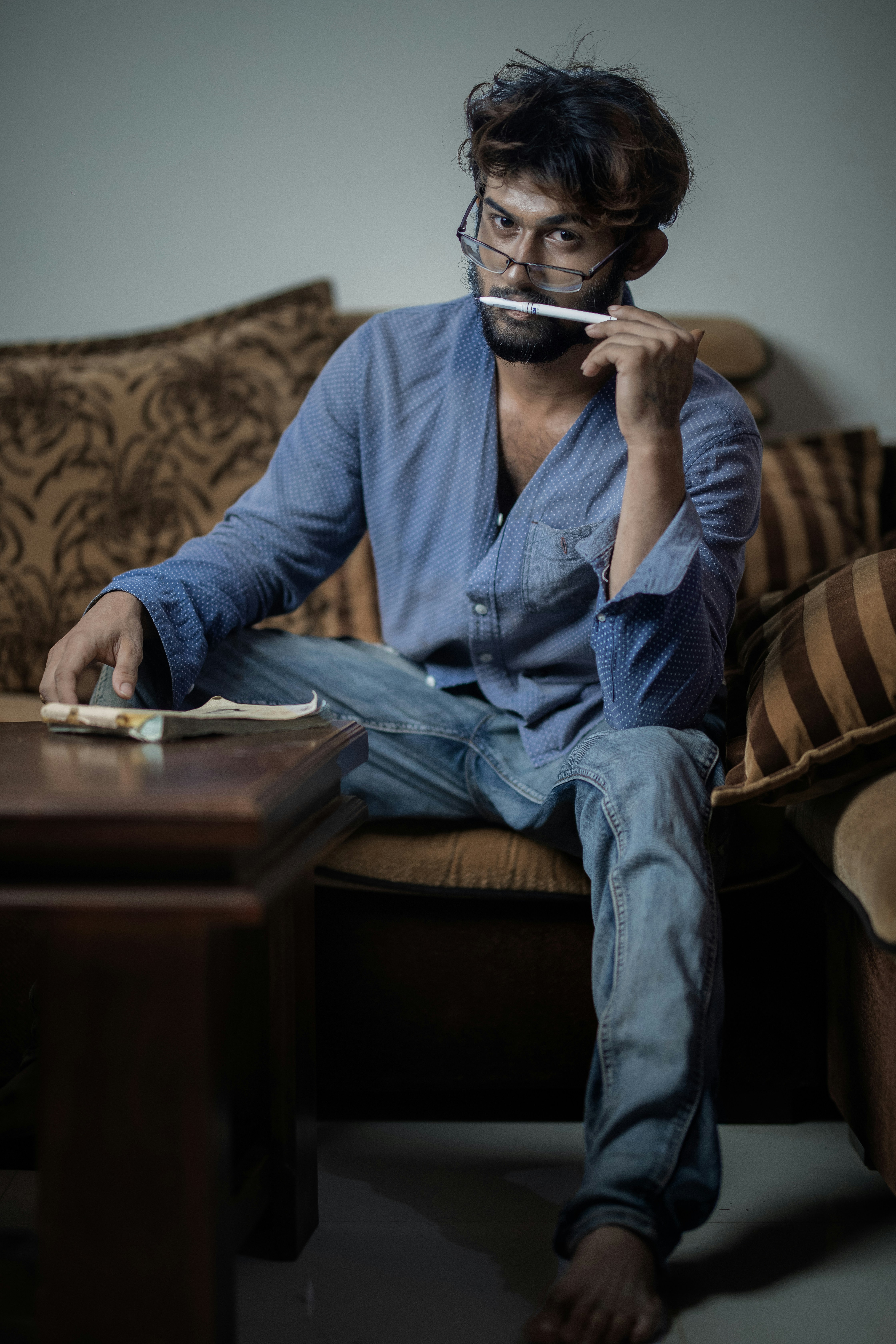 Man with glasses on couch, pen in mouth, looking thoughtful.