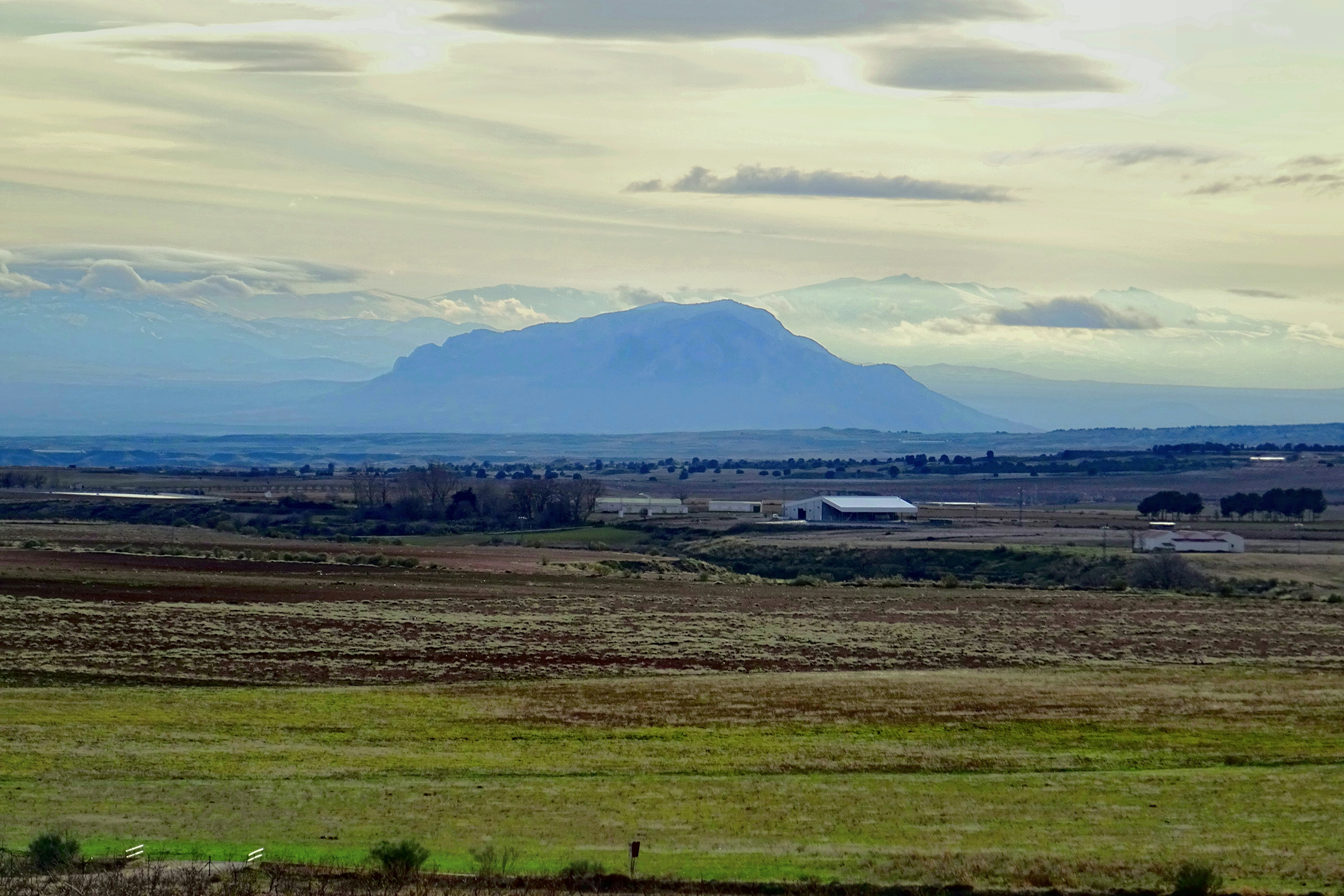 Wide rural landscape with distant volcano-like mountain on the horizon and flat farmland in foreground; a soft, overcast sky suggesting late afternoon light.