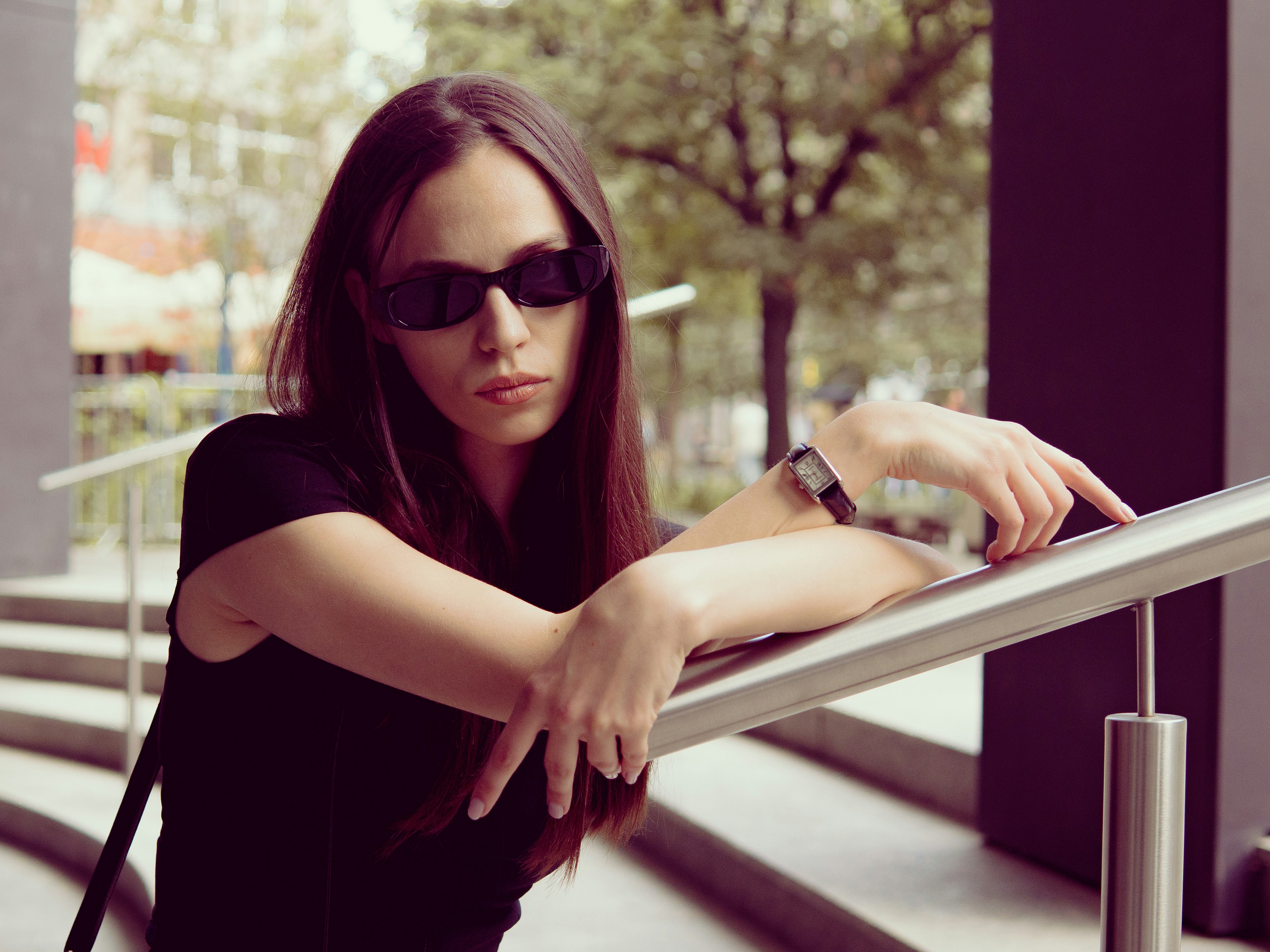 Dark-haired woman in sunglasses leans on a railing in an urban setting, captured with a moody, cinematic tone. The scene emphasizes attitude and composure with a shallow depth of field.