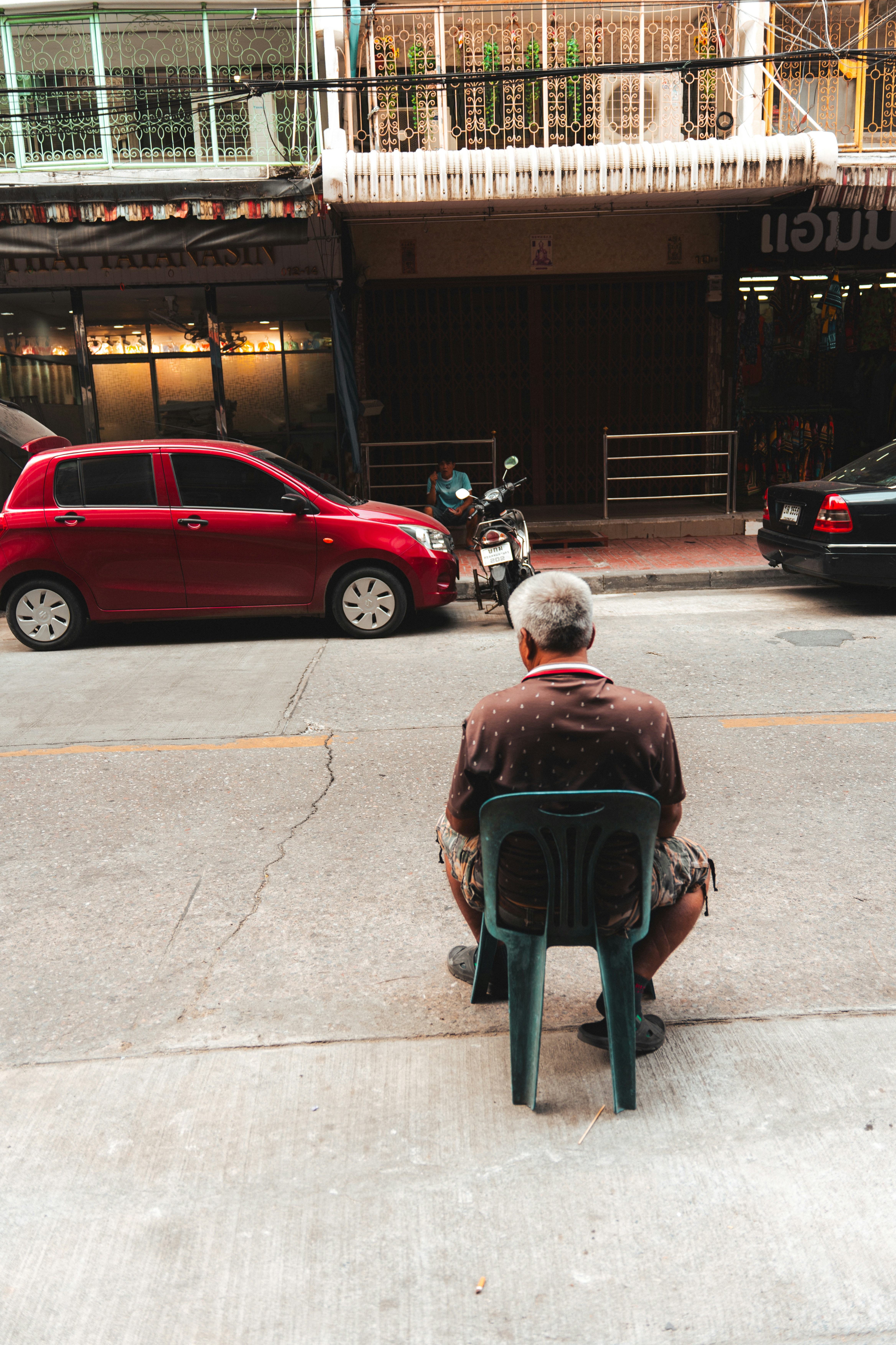 Rear View of a Street Elder Seated on a Green Chair facing an urban storefront