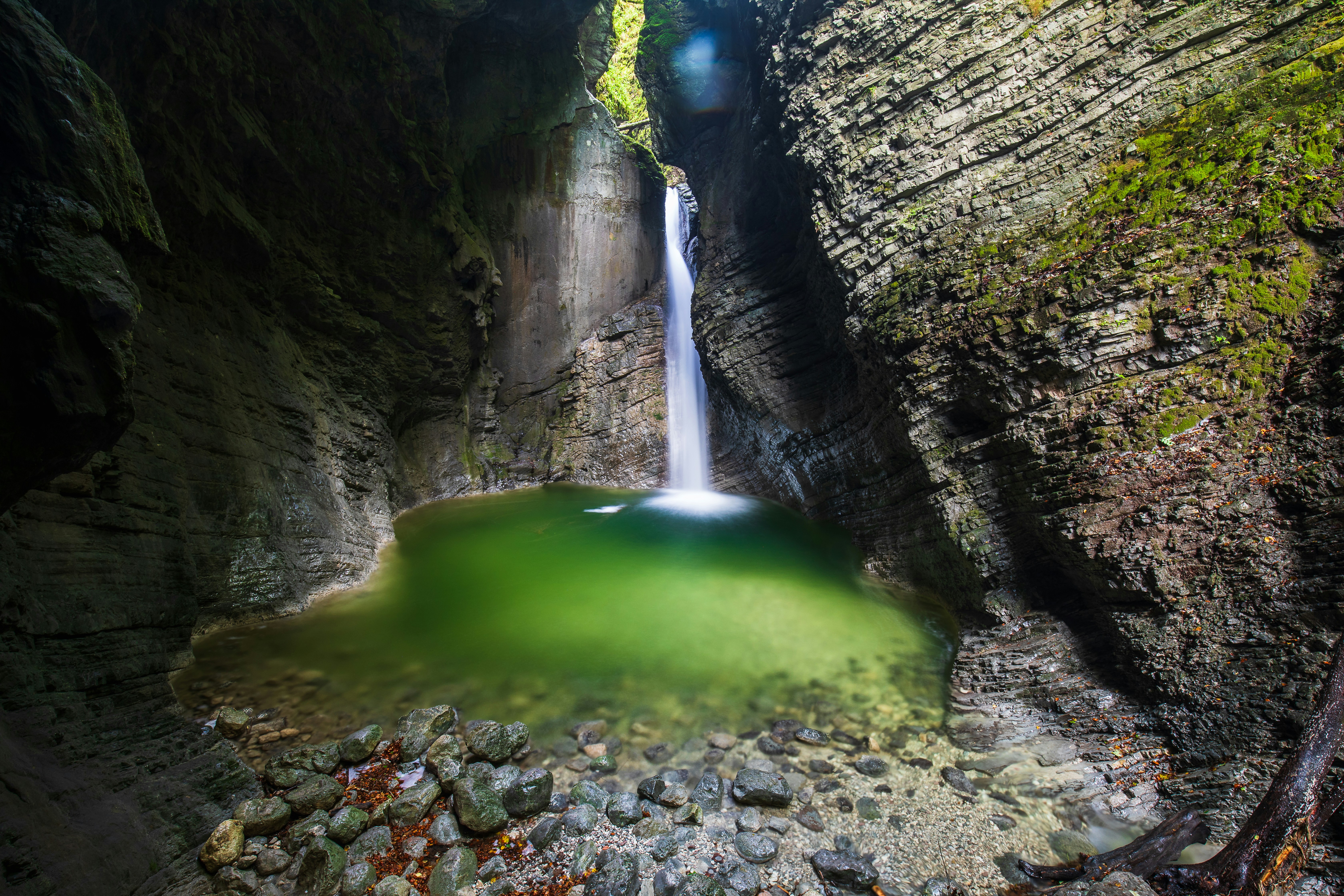 A mossy gorge with a slender waterfall feeding a vivid green pool, framed by rugged rock walls and a rocky shoreline.