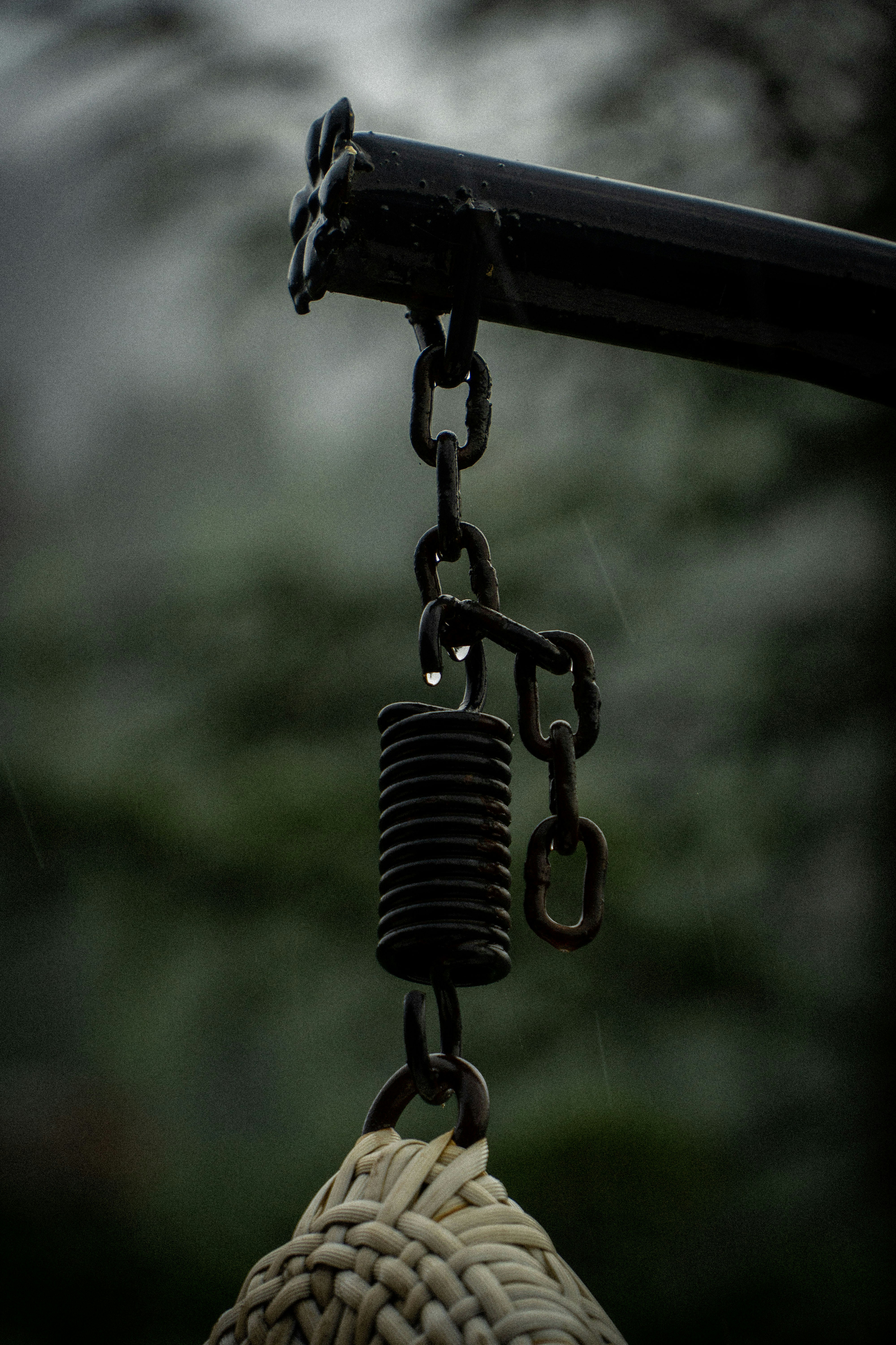 Close-up of a metal chain and spring hanging from a bar, with blurred natural background and subtle raindrops. It highlights industrial detail and texture in muted tones.