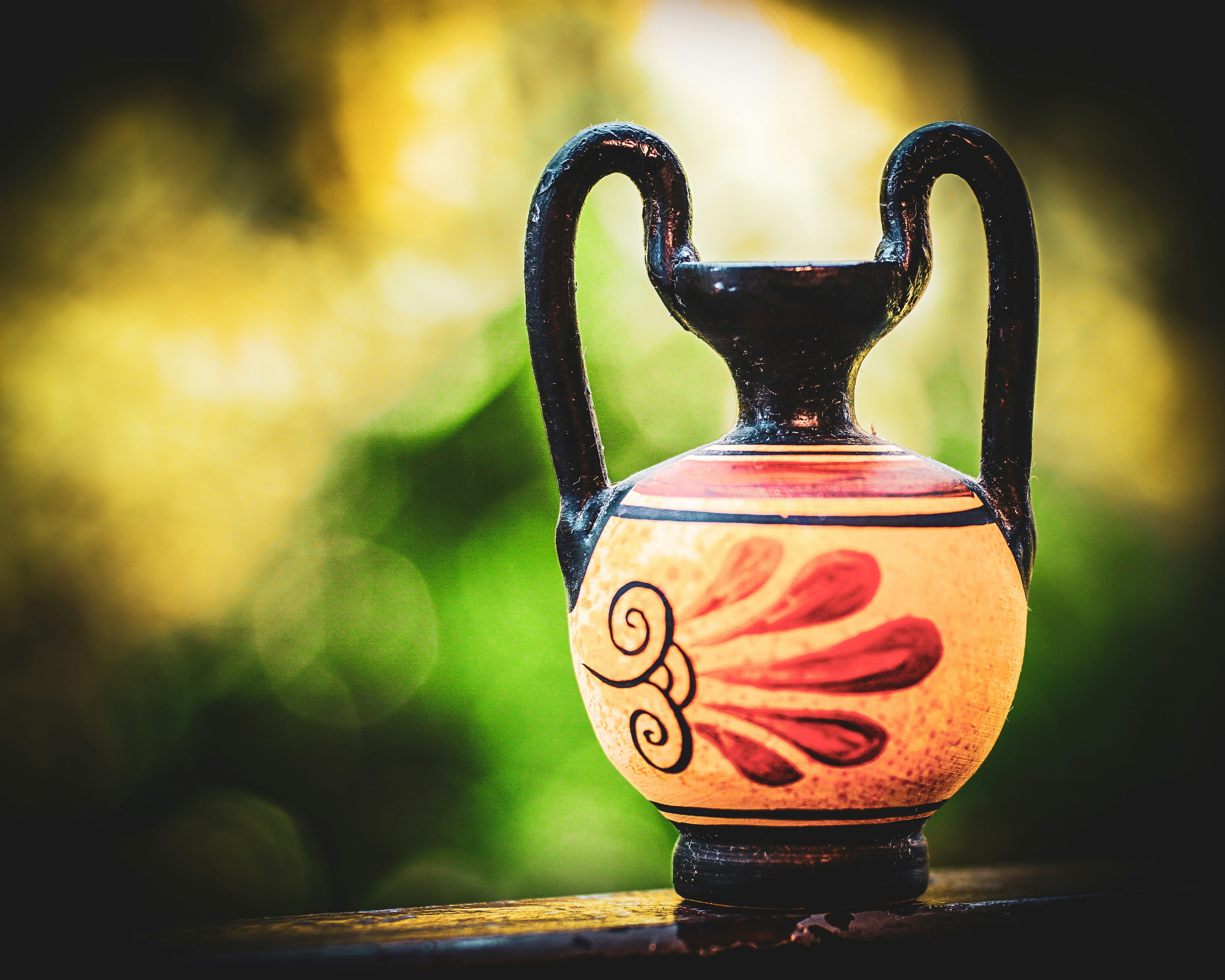 A decorative ceramic vase with two handles sits on a ledge, bathed in warm sunlight with a soft bokeh-green background, emphasizing the painted floral motif. The scene highlights craft detail and color texture.