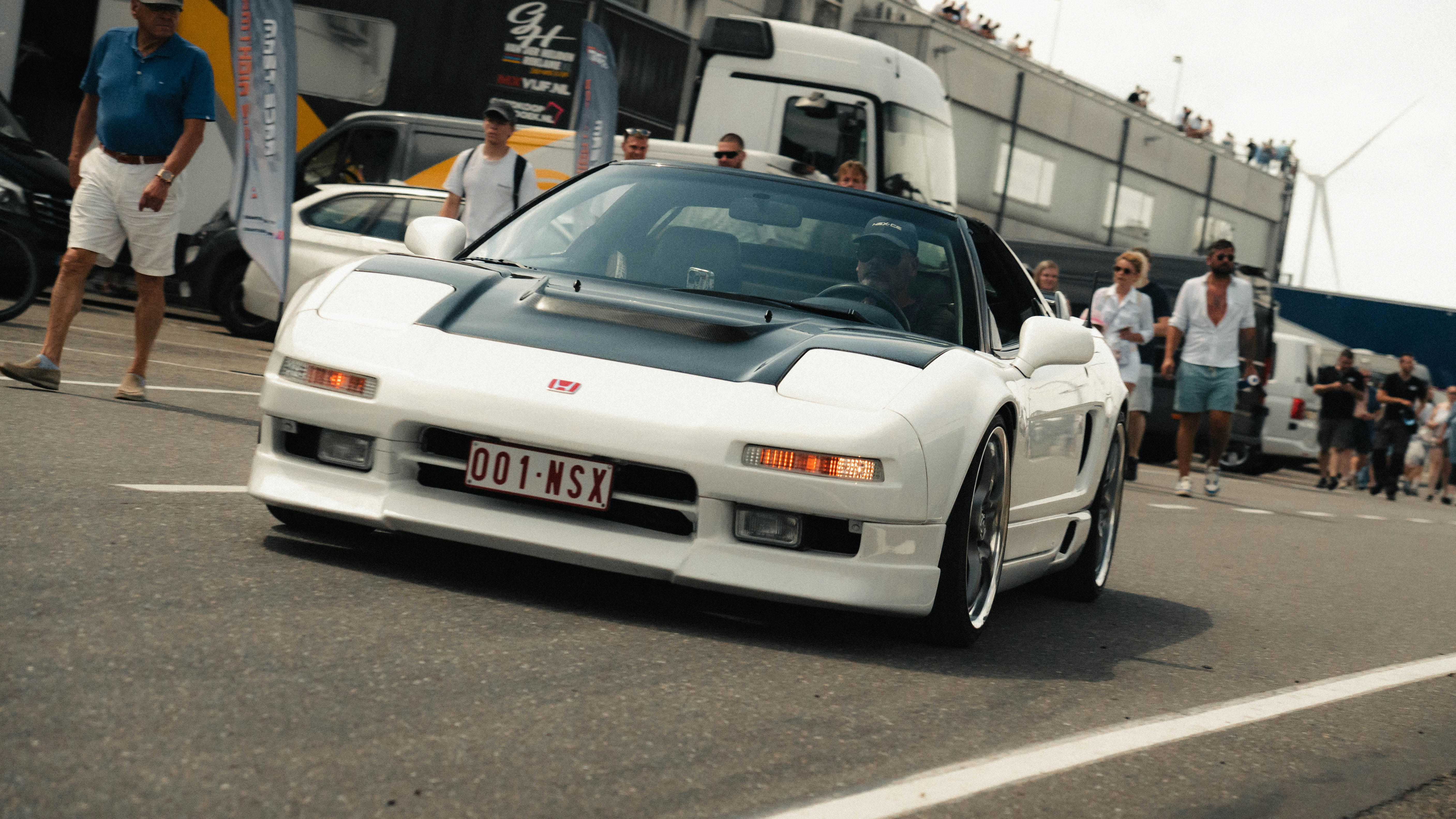 White Ferrari sports car with open-top driving past a crowd at a race event; logos, trailers and spectators line the background; high-energy street-style capture.