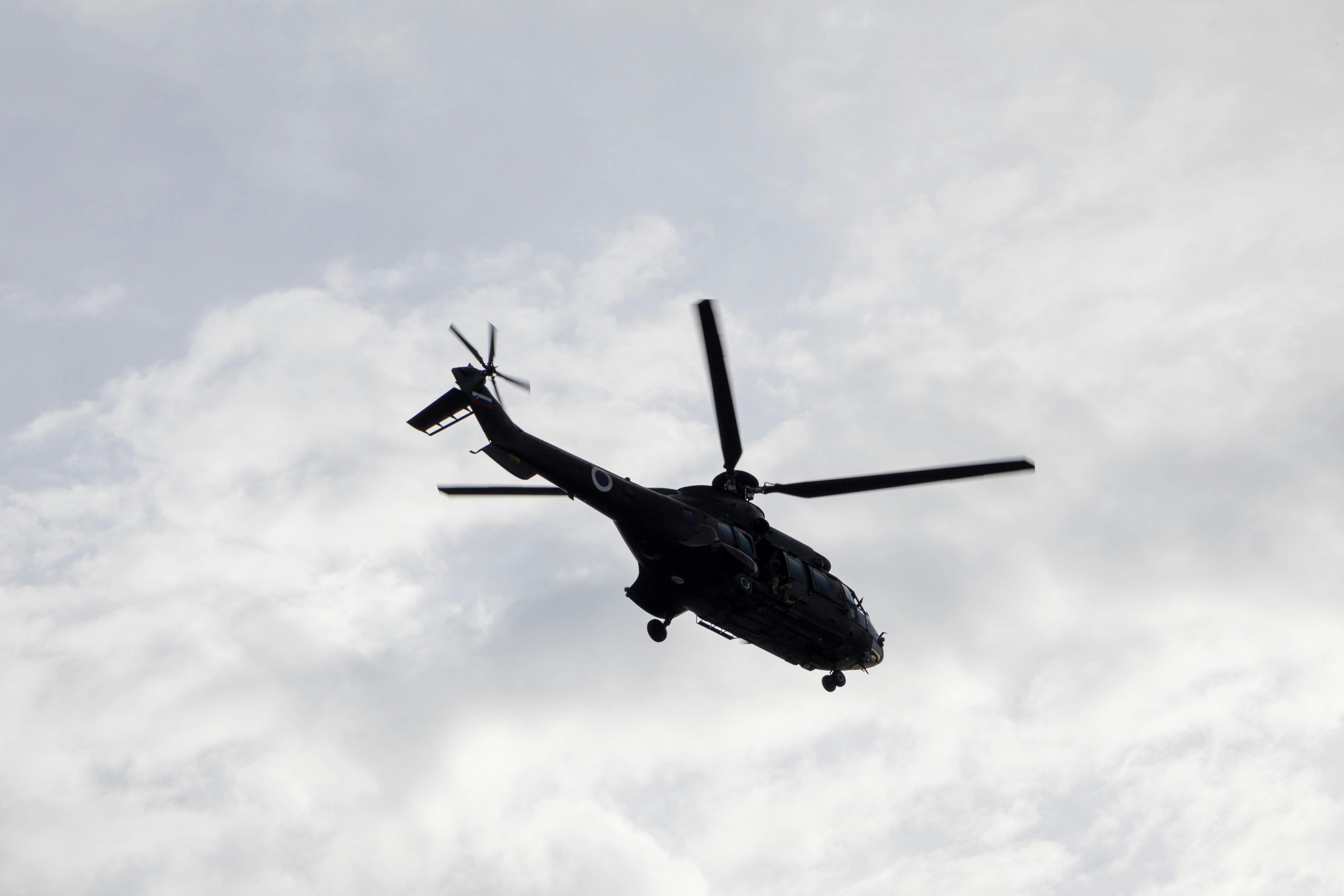 A dark helicopter flies against a cloudy sky.