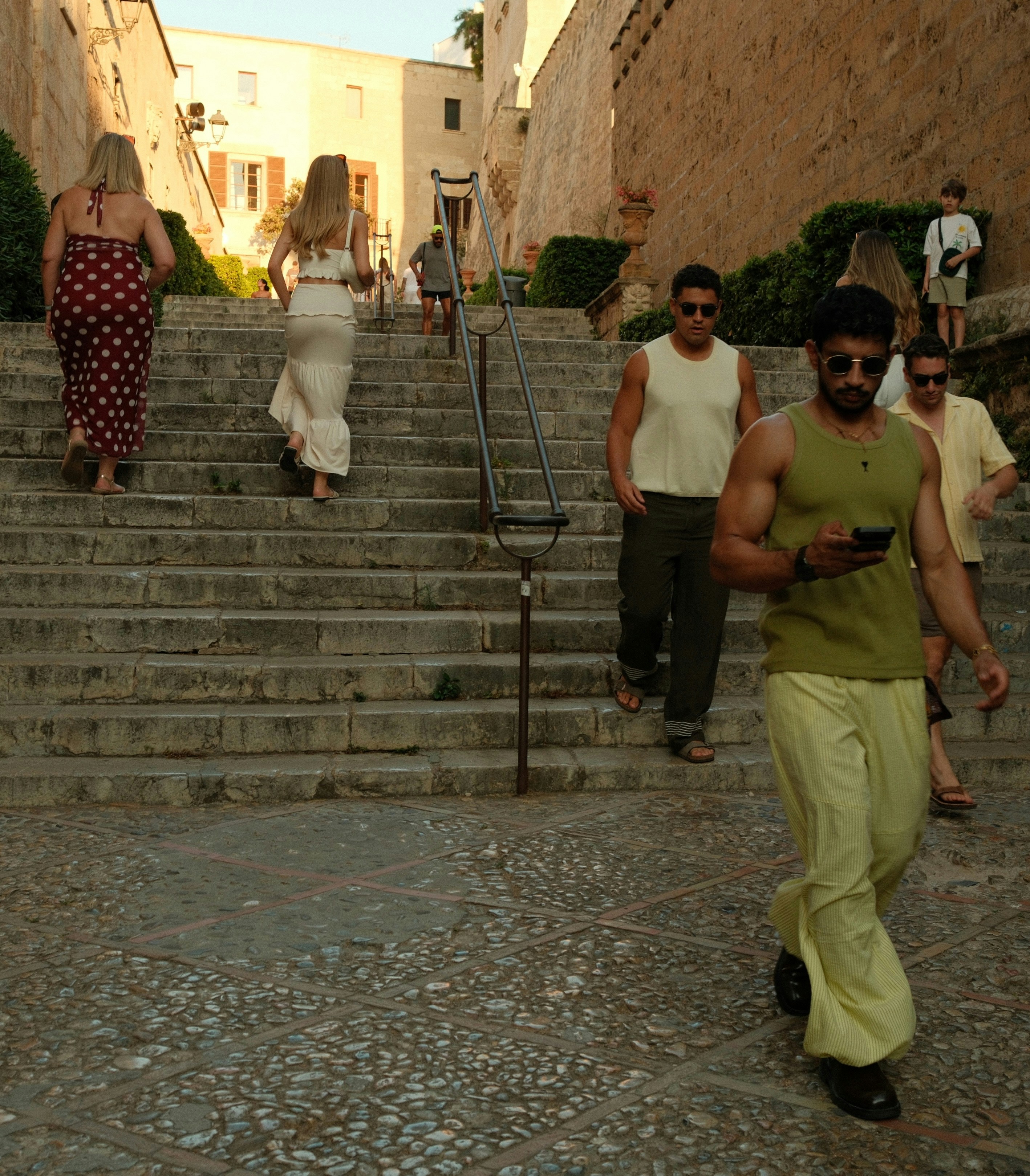 Dramatic scene on sunlit stone stairs with pedestrians moving through a warm European plaza; a foreground man in green sleeveless shirt checks his phone while others ascend and descend, revealing textured cobblestones and stone walls.