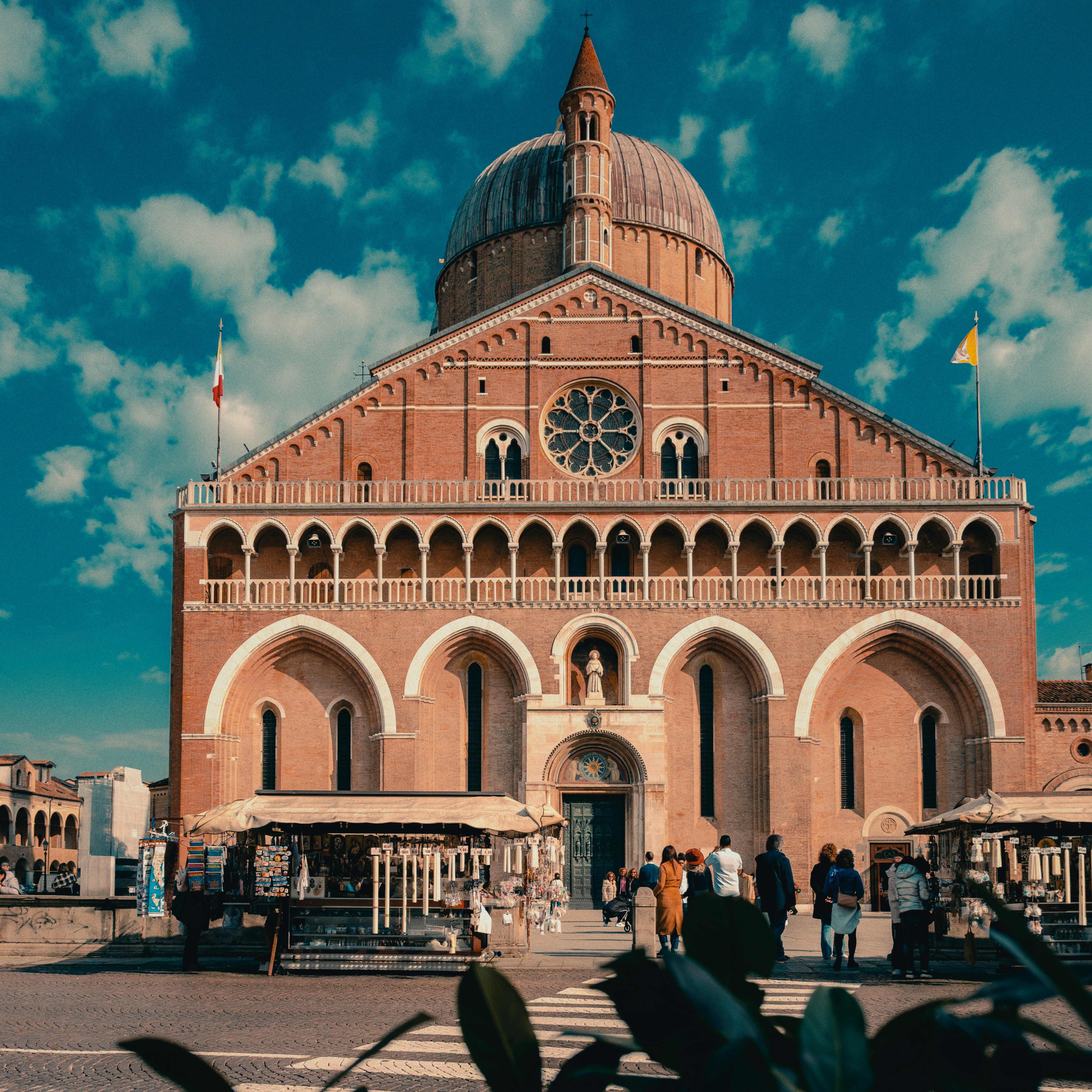 A brick basilica with a large central dome and arched facade stands under a bright blue sky, with people and market stalls in the foreground.