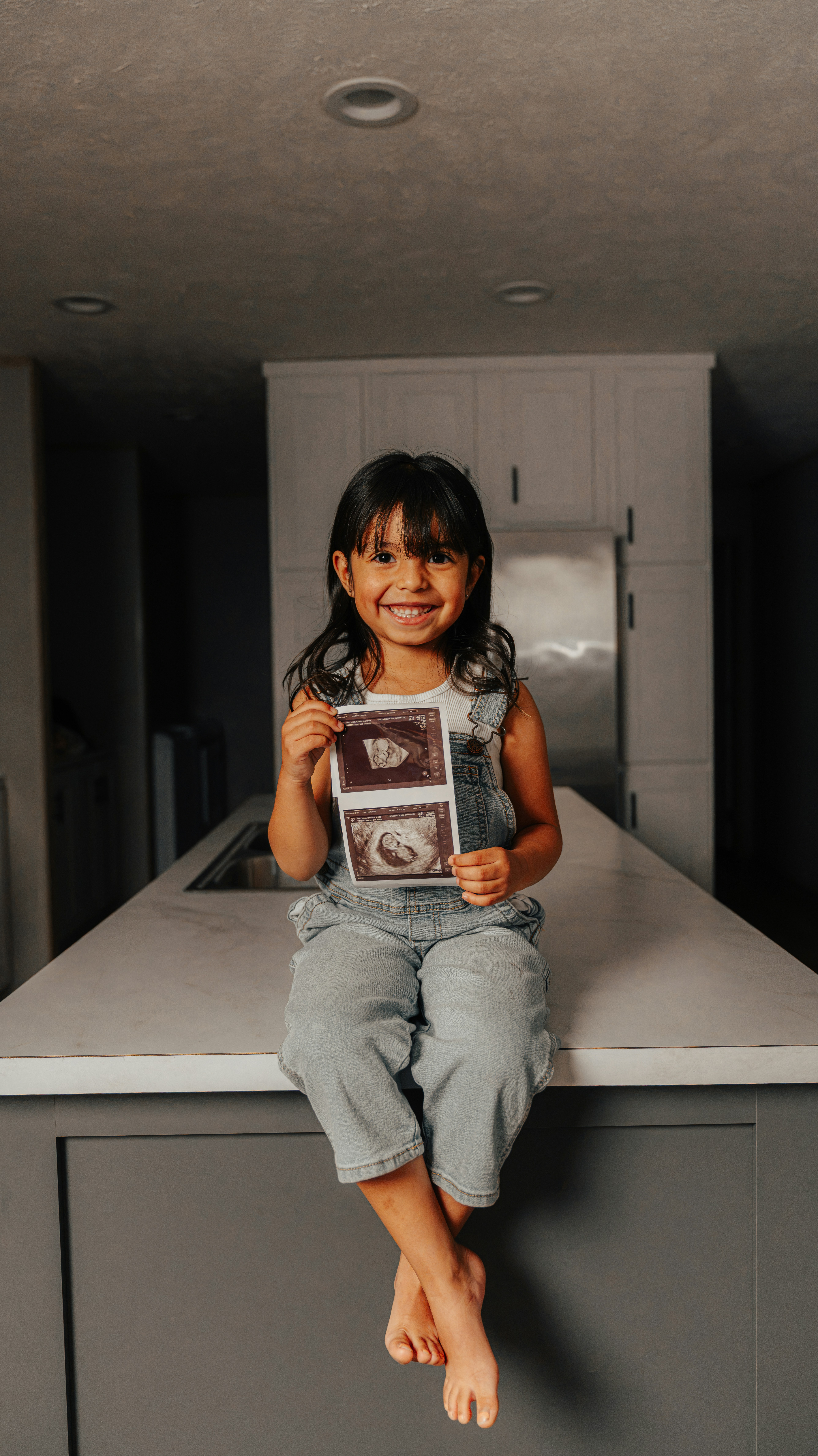 Smiling child sits cross-legged on a kitchen island, holding ultrasound photos amid a modern, softly lit backdrop.