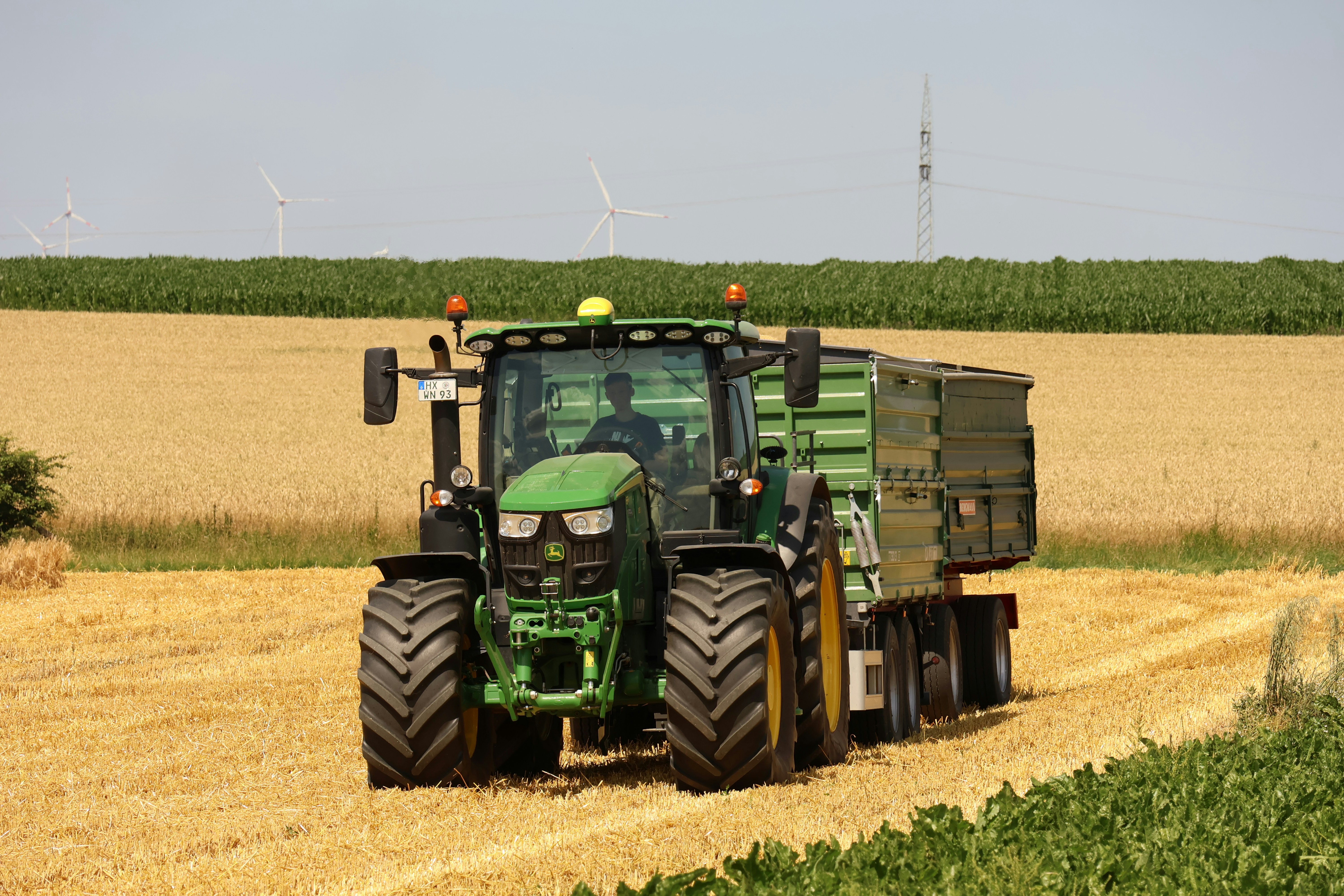 Green tractor pulling a trailer moves through a sunlit harvested field with a distant row of crops on the horizon and wind turbines in the background.