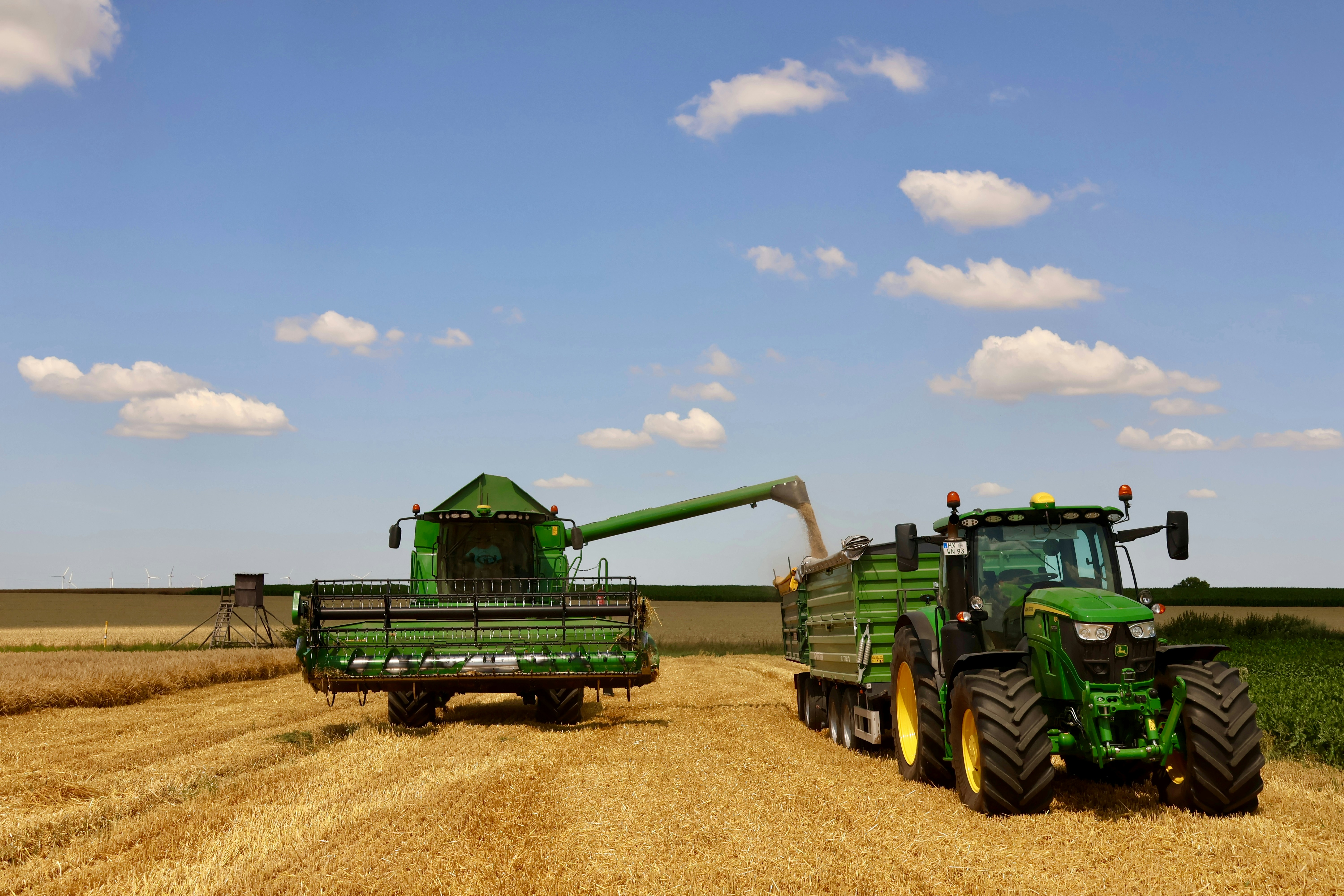 A pair of green agricultural machines sit in a harvested field under a bright blue sky, ready to continue the work. This is a straightforward agricultural scene with a combine harvester and a tractor in sharp focus.