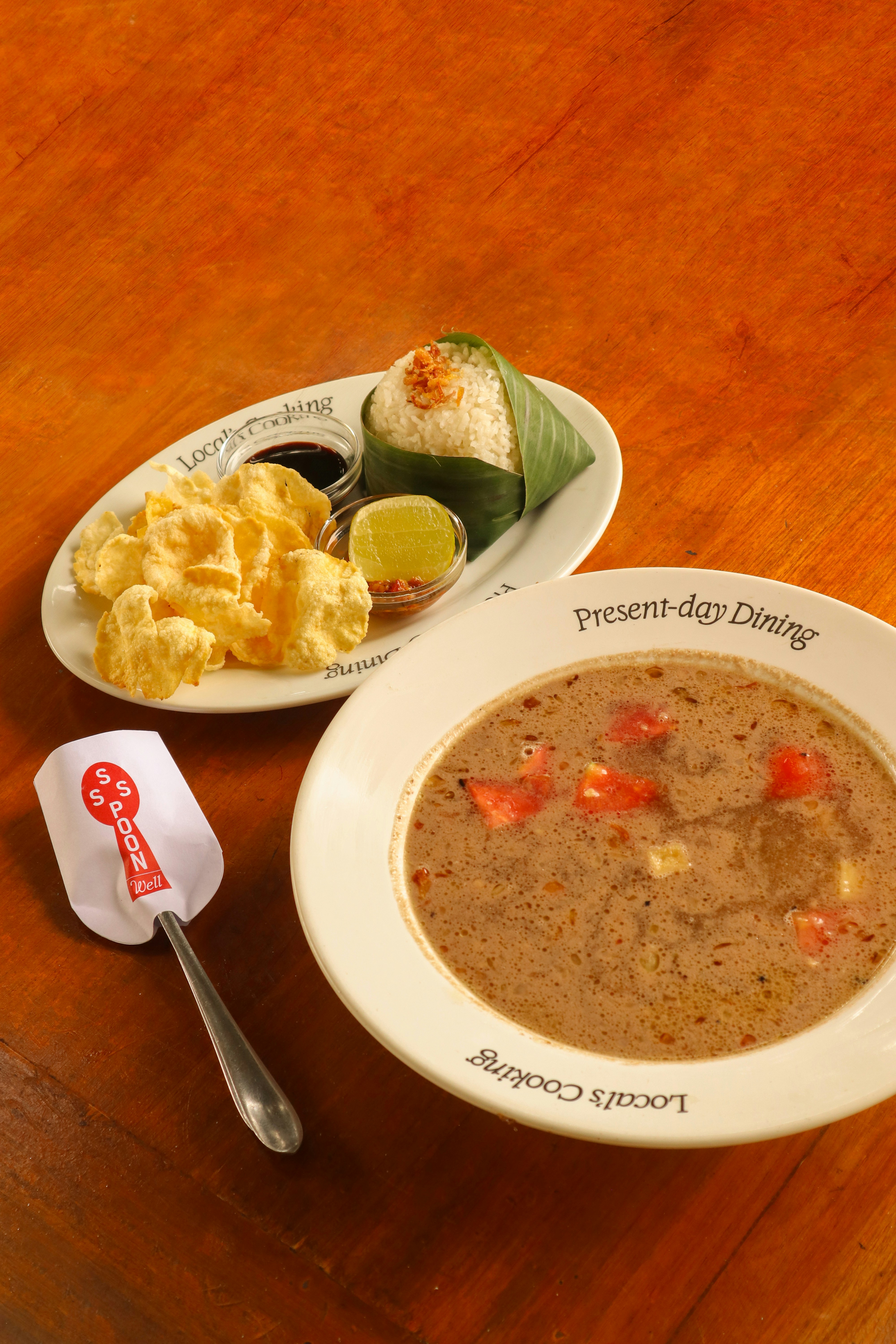 A bowl of creamy soup with tomatoes sits near a plate of crispy chips and a wrapped rice dumpling on a warm wooden table; warm tones and casual plating evoke a cosy dining moment. Note: plate and spoon indicating a casual restaurant setting.