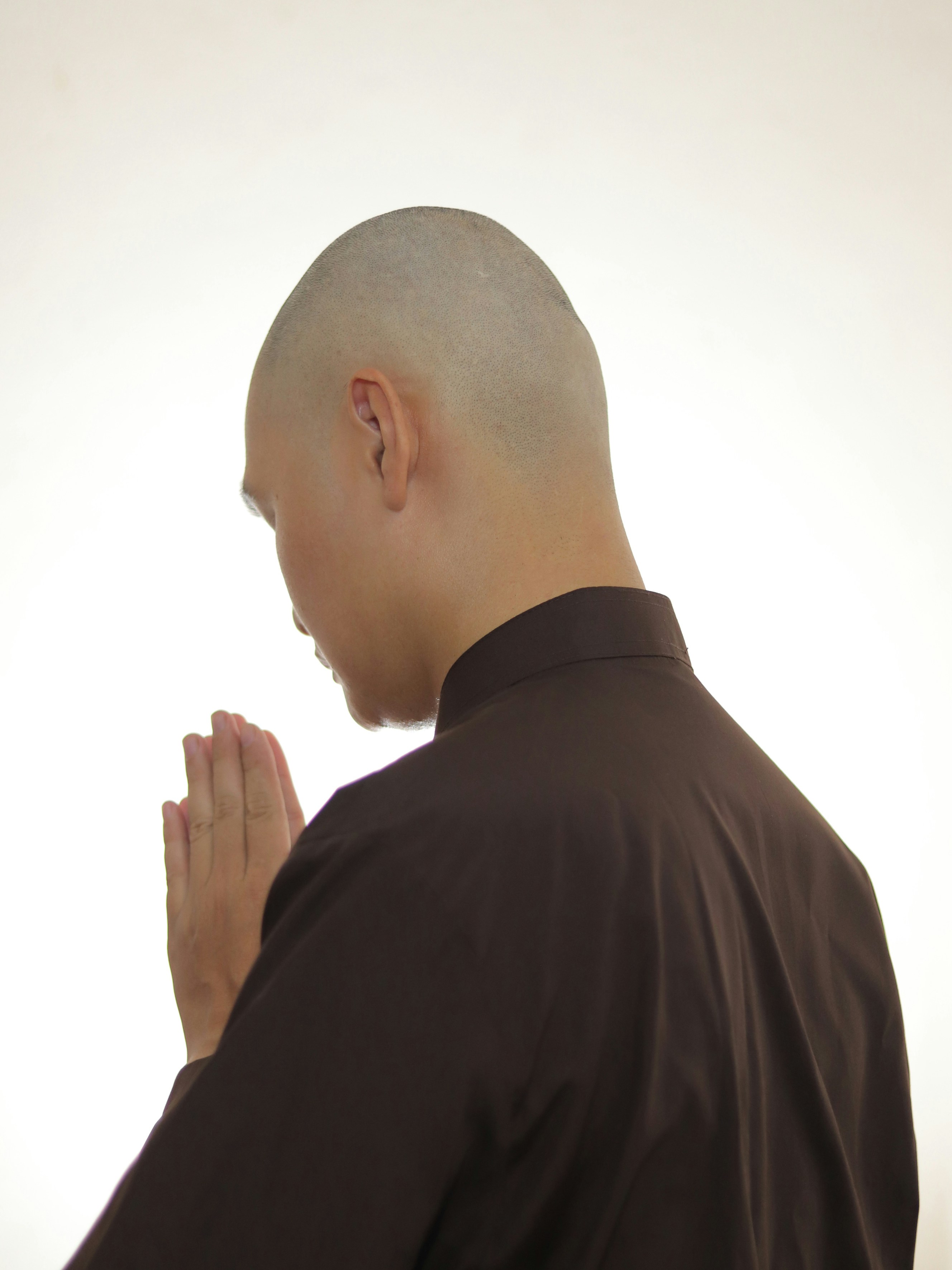 Minimalist side-profile of a man with hands pressed together in prayer, set against a soft, bright background. A studio portrait emphasizing posture and calm expression.