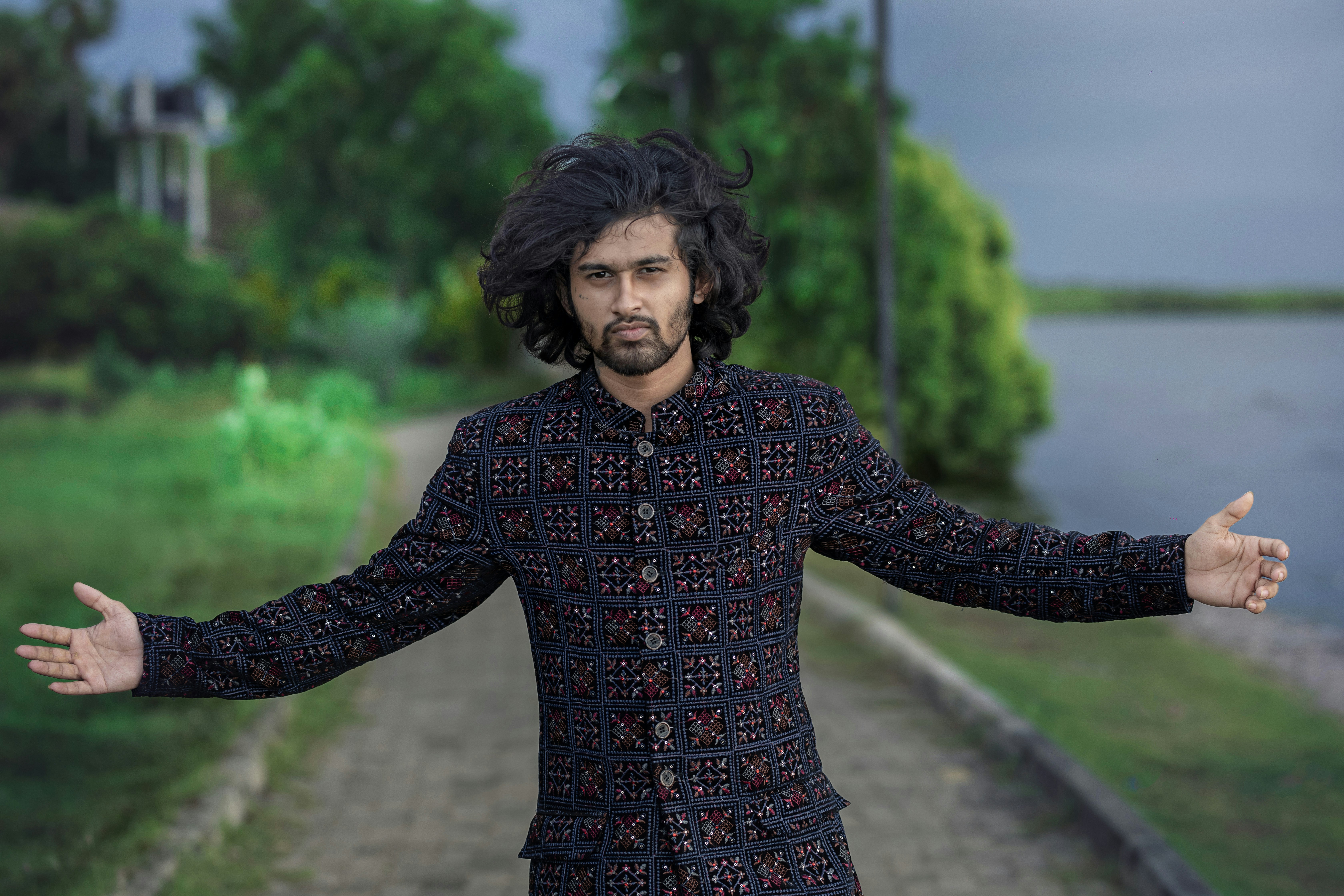 A stylish outdoor fashion portrait of a young man standing by the waterfront, wearing a dark embroidered designer jacket with intricate geometric patterns. His windswept hair and well-groomed beard add a bold, modern look, while the calm water and soft natural light create a cinematic atmosphere. The subject is looking away from the camera, giving the portrait a confident and contemplative feel with a shallow depth of field that beautifully separates him from the background.