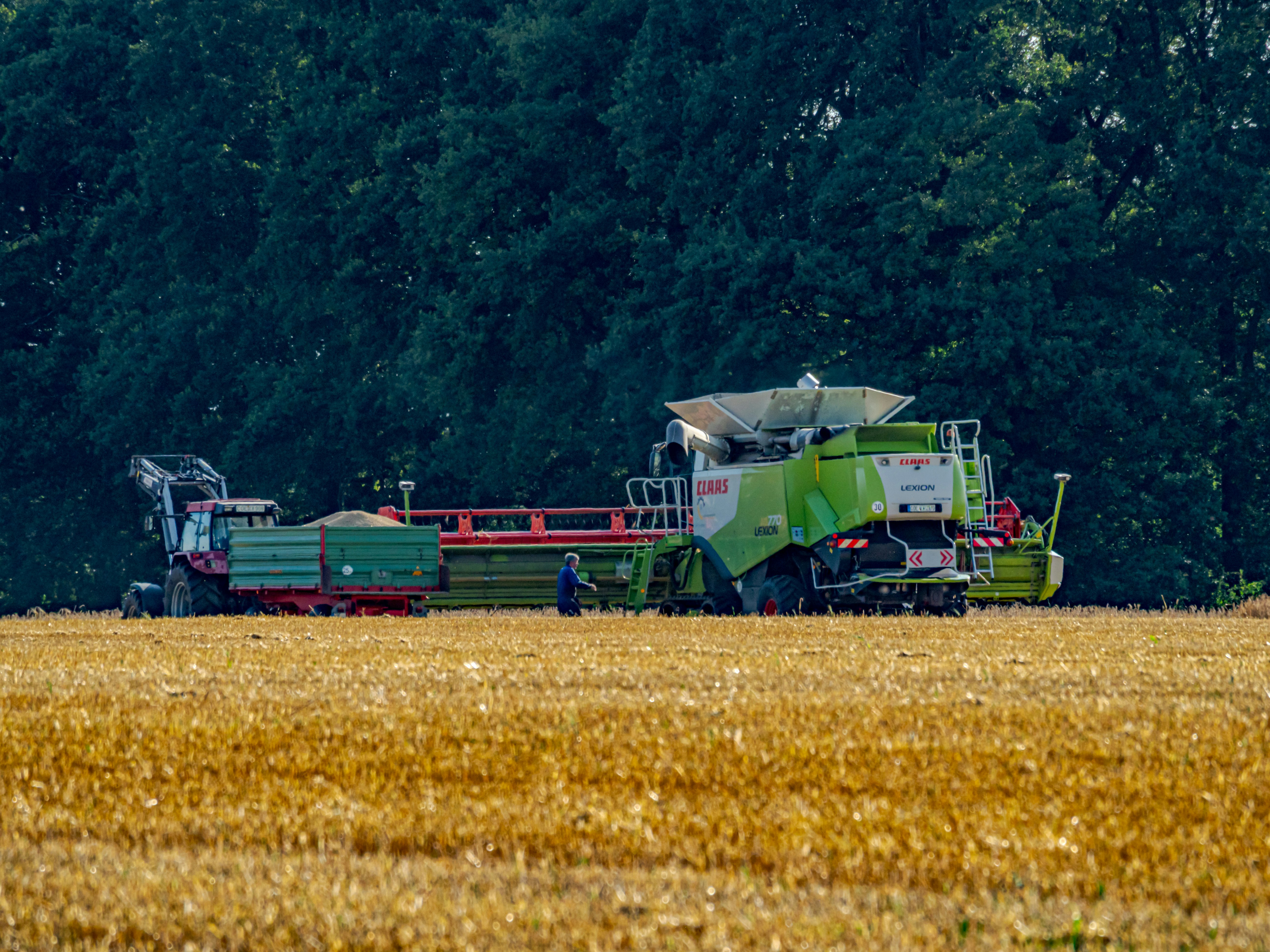 Golden-stubbled field with a bright-green harvester and adjacent machinery set against a dark tree line; a distant operator stands by, highlighting modern harvesting equipment in use.