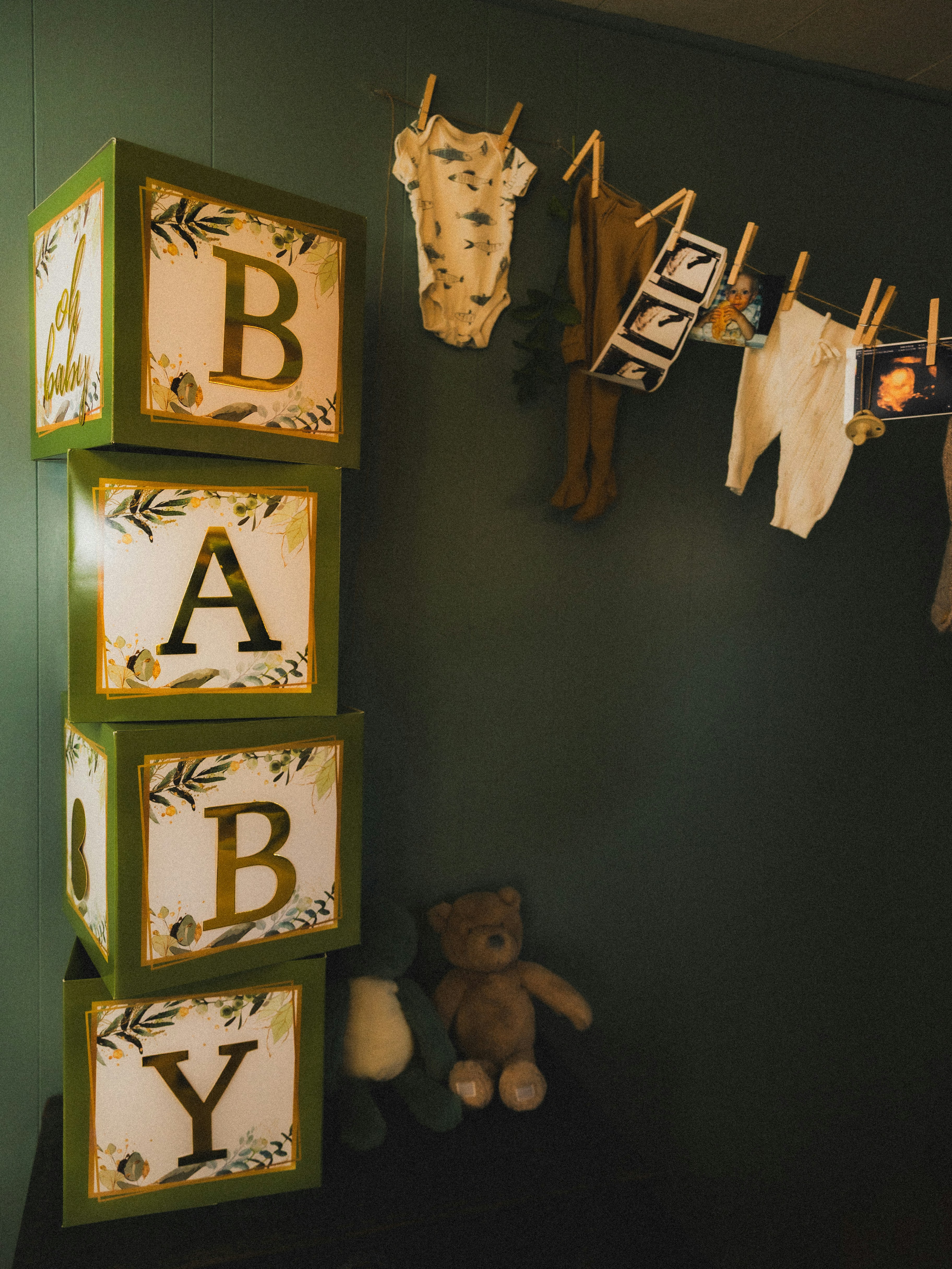 Stack of green-framed blocks spelling 'BABY' sits beside a dark wall; clothesline with photos and small garments drapes above, with stuffed toys at the base. A warm, intimate nursery vignette.