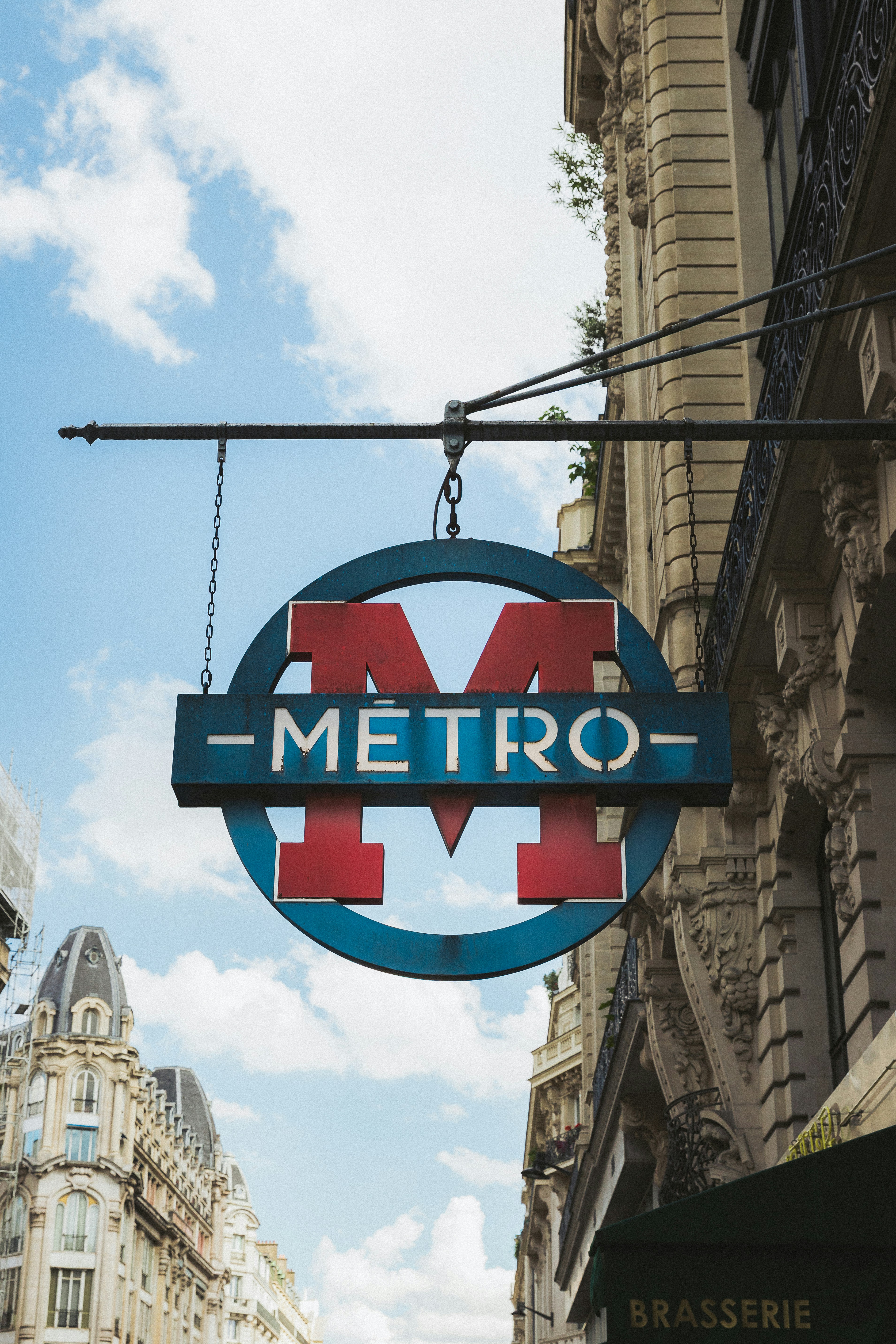 A classic blue-and-red Metro sign hanging over a Parisian street, framed by ornate building façades and a bright blue sky with clouds. This is a photograph of a public transit emblem capturing urban architecture and signage.