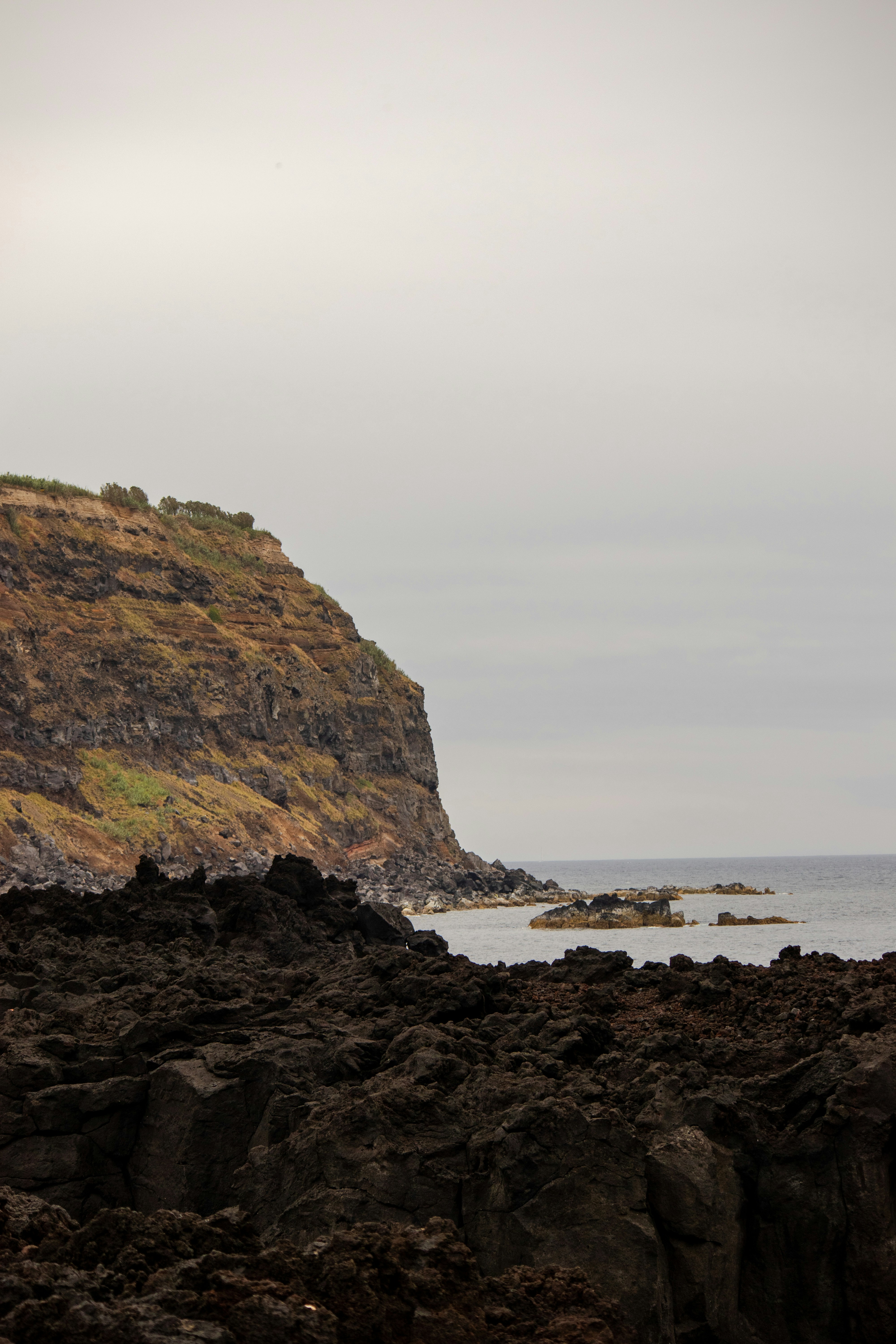 Rugged volcanic shoreline with a towering cliff meeting a calm sea under soft, overcast light. The foreground is dominated by dark lava rocks leading to a distant promontory.