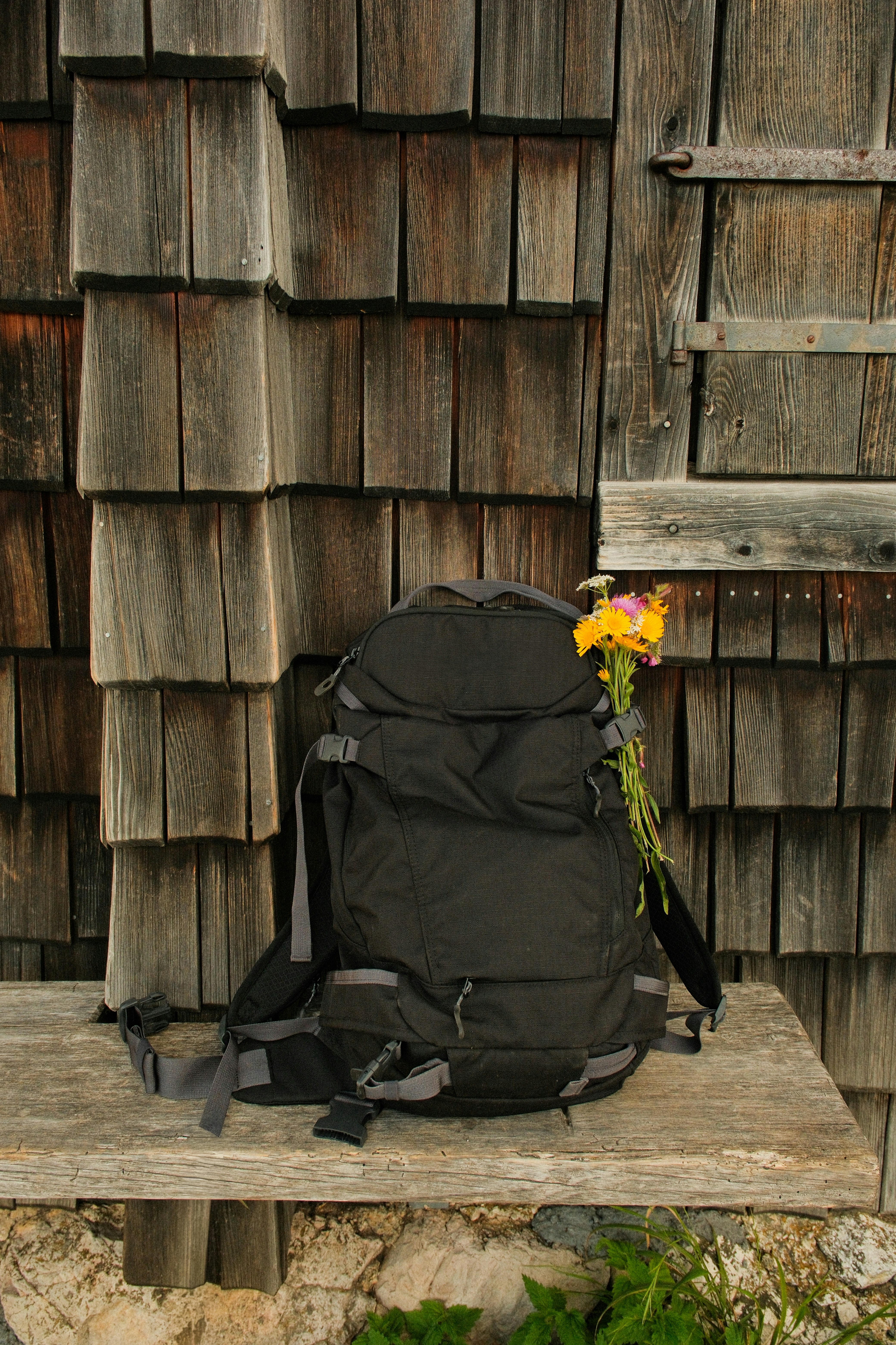 Backpack resting on a weathered wooden bench against a rustic door; a small bouquet tucked into the side strap adds a pop of color. An outdoor scene with natural textures and muted tones.