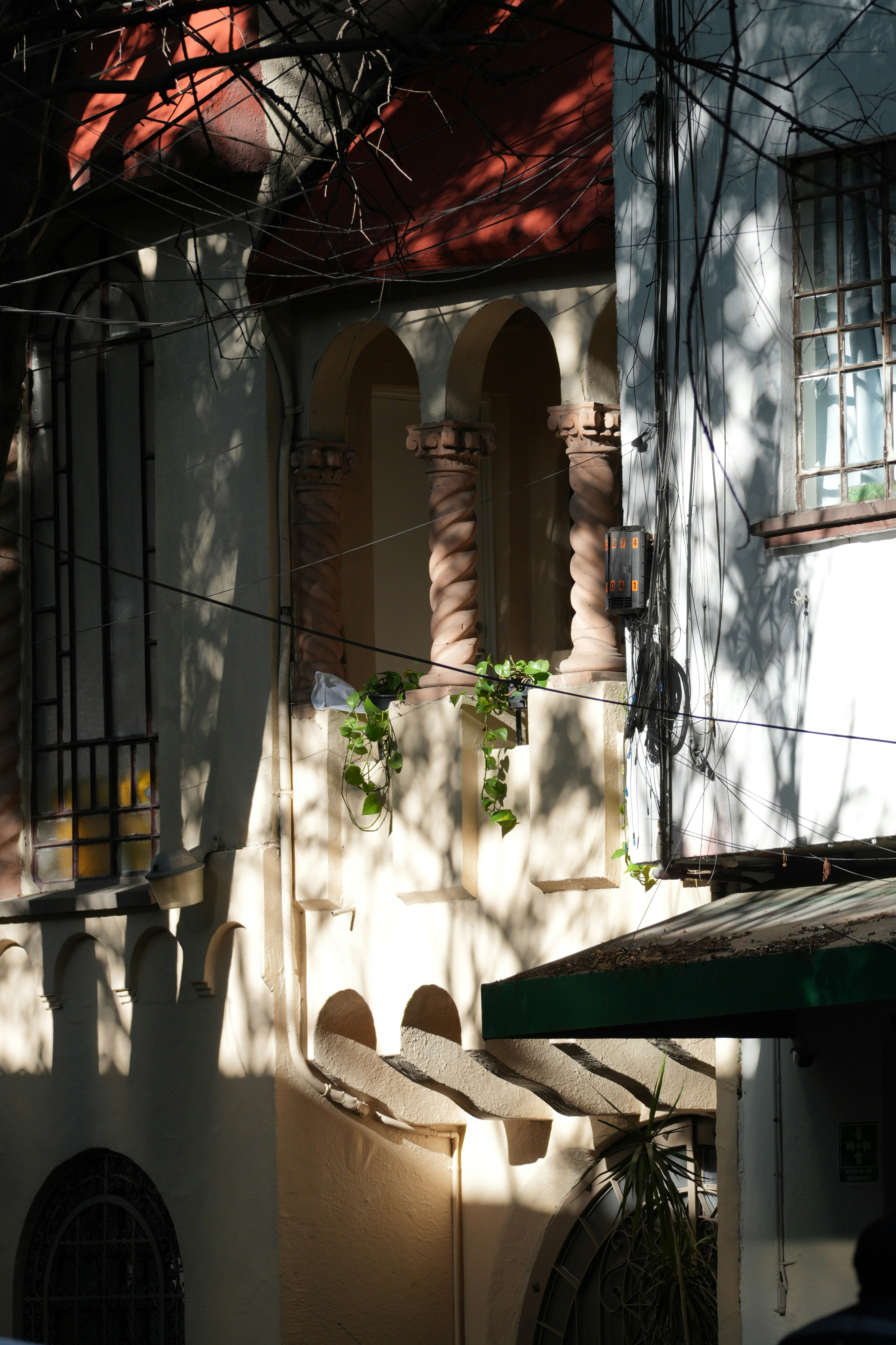 Sunlit façade with decorative balcony columns and hanging vines; architectural details and shadows create contrast on a shaded street scene.