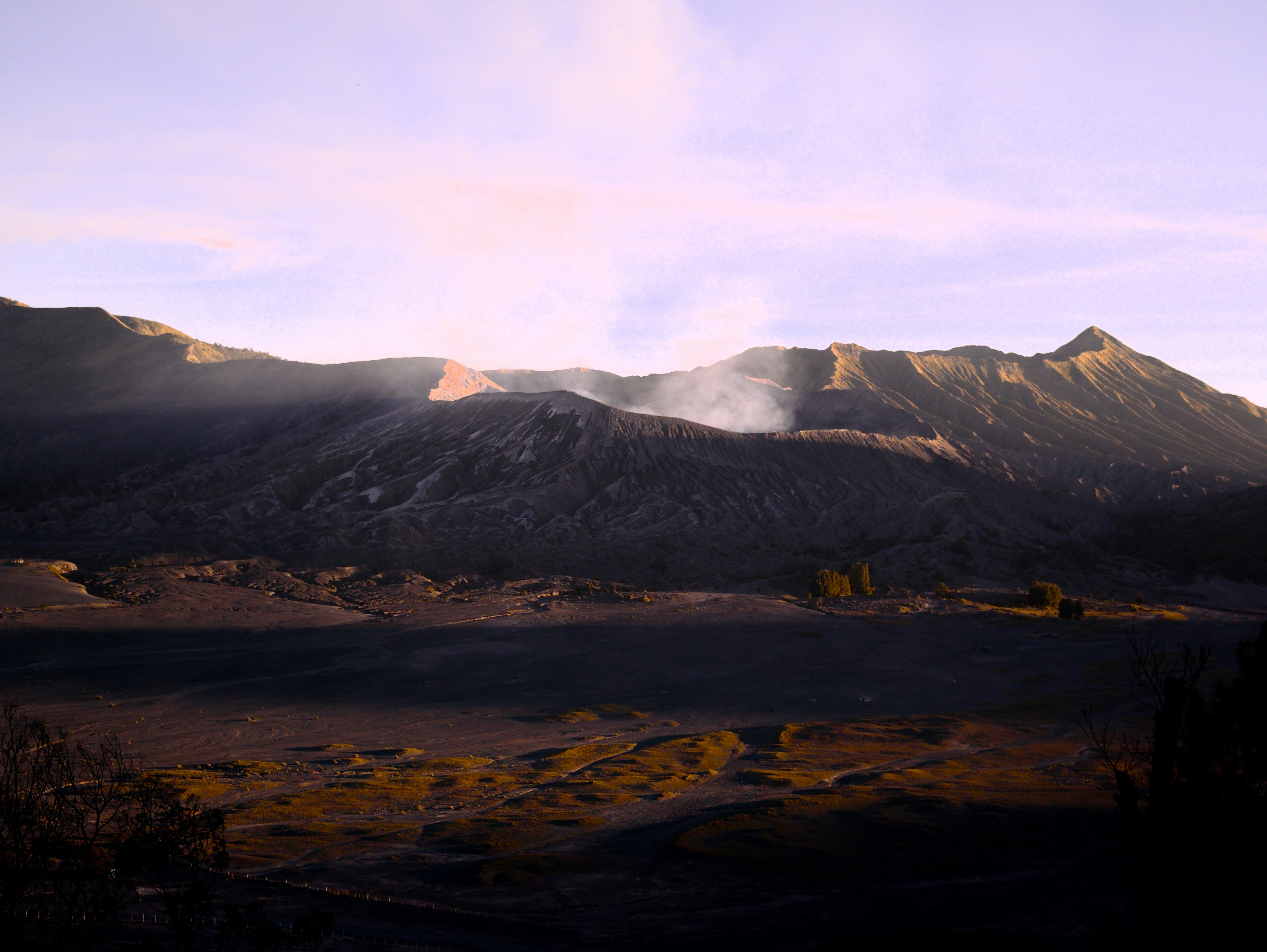 Lava field with a serrated ridge at dusk, wisps of steam rising from the crater and a pale pink sky in the background.