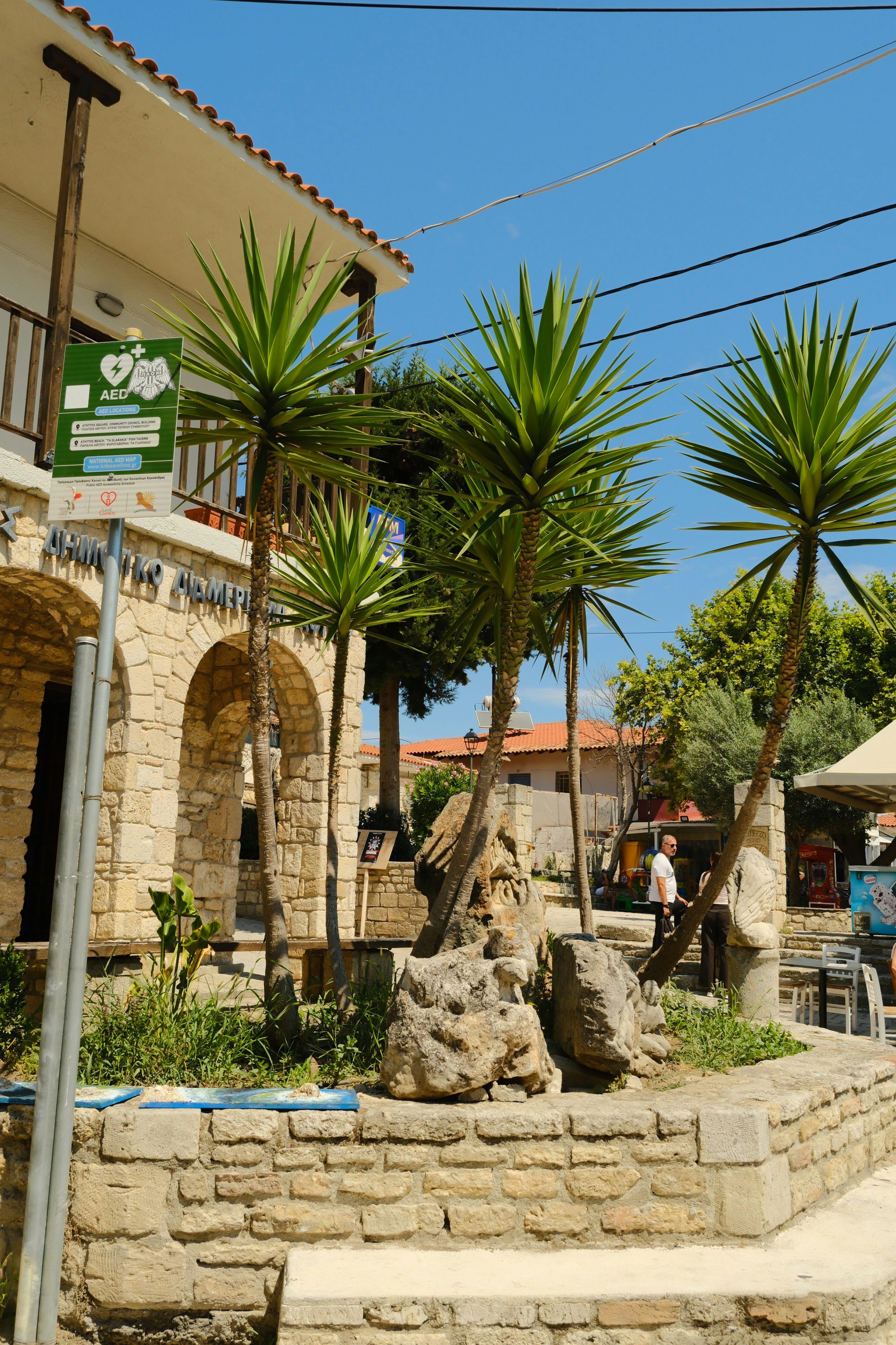 Desert Courtyard in Sunlit Stone: Palm Trees, Café, and Aed Sign