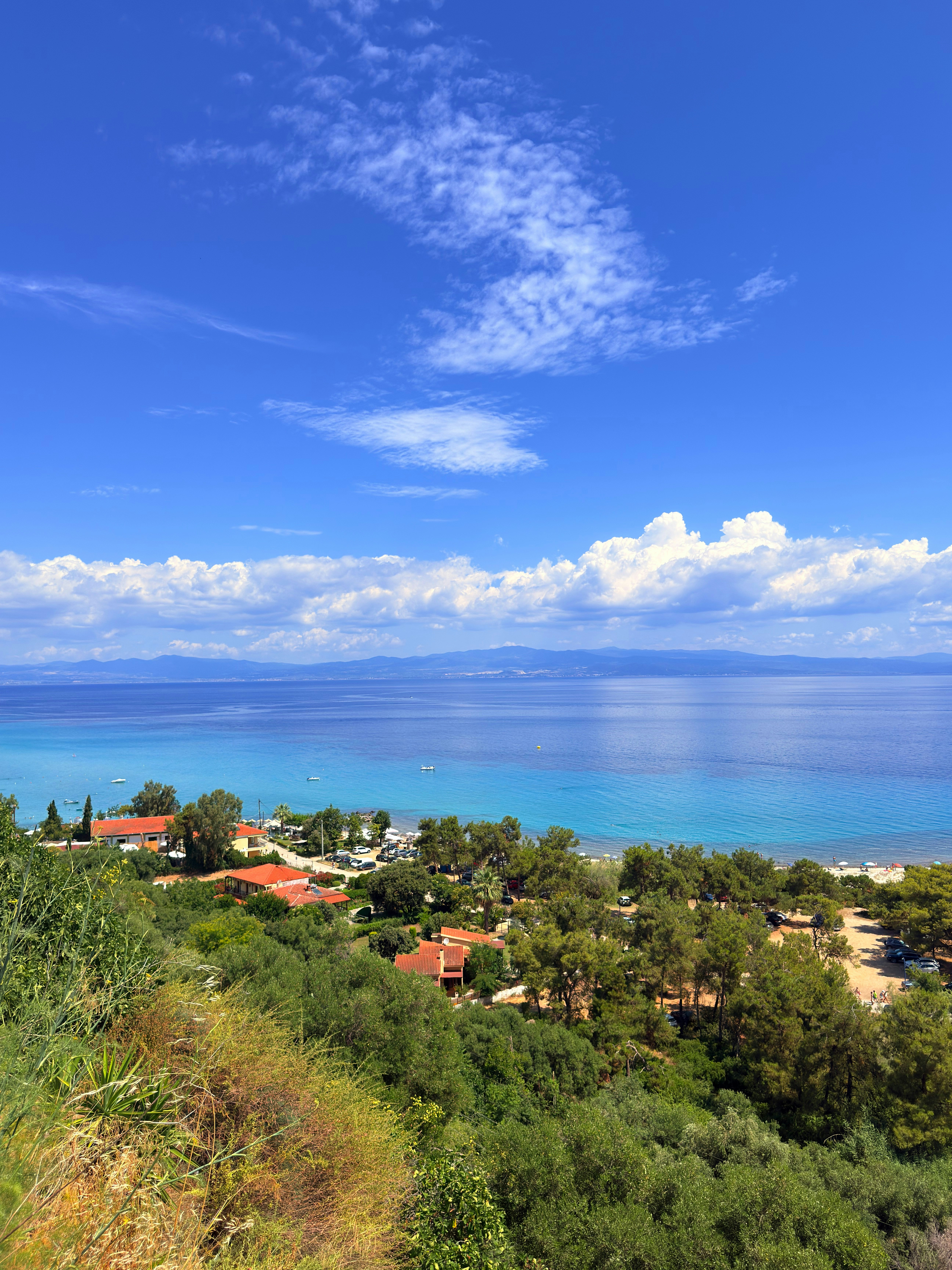Sunlit coastline with turquoise sea, sandy beach, and clustered coastal homes below a bright blue sky.