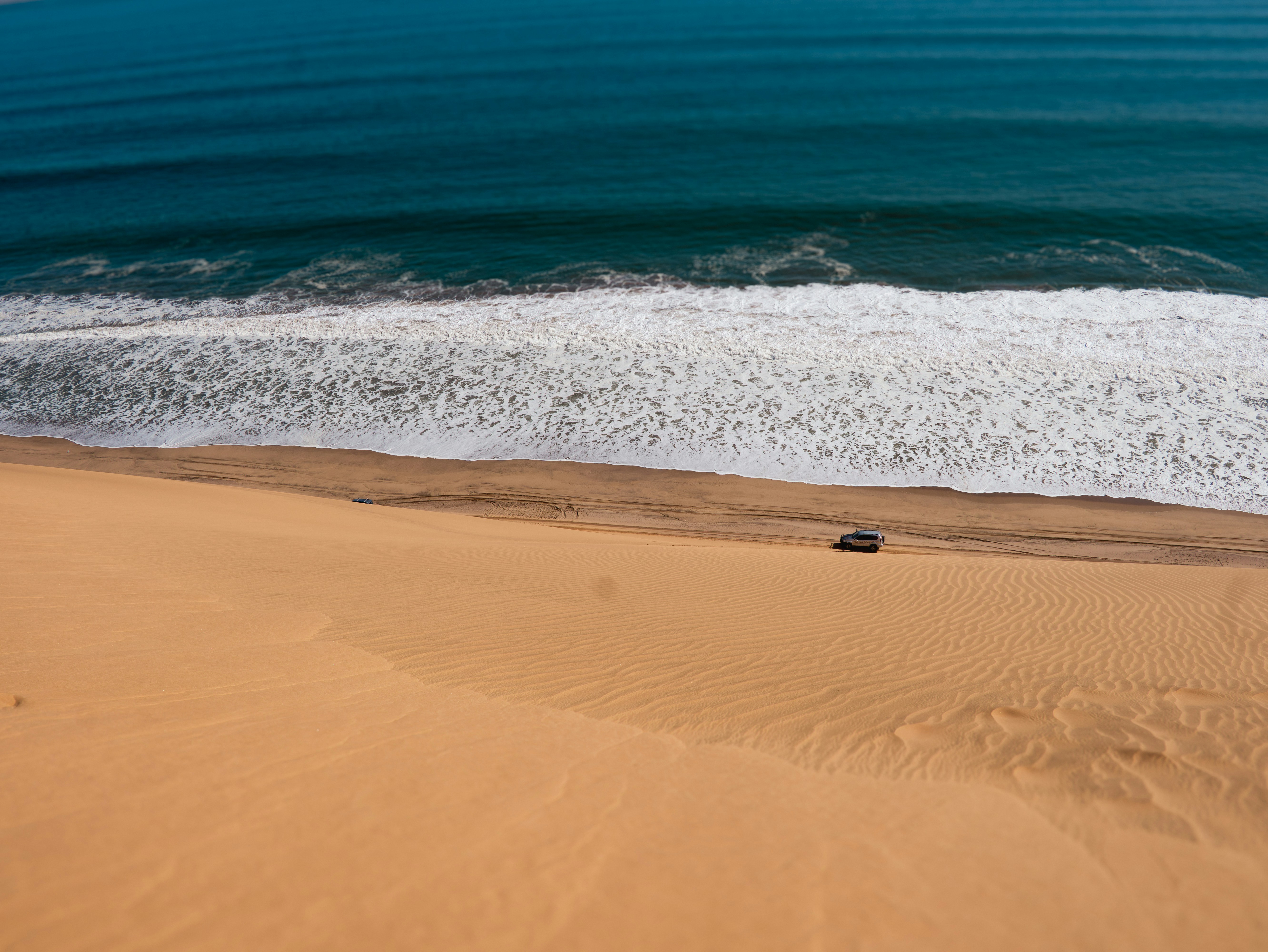 Car on a sunlit sandy beach with a rolling ocean behind, waves crashing near a distant shoreline. A minimal scene emphasizing vast dunes and open horizon.