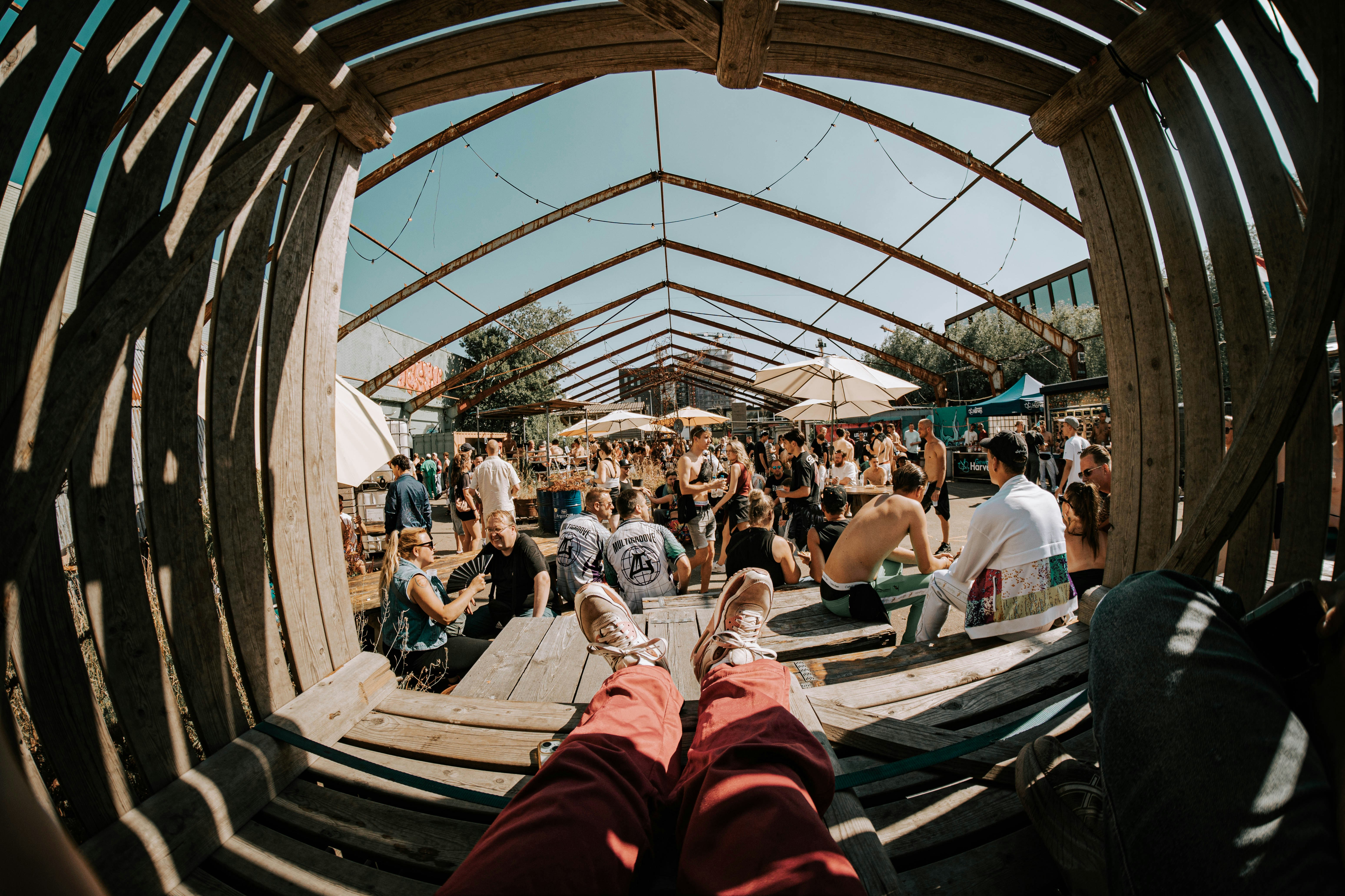 People mingle beneath a wooden arched structure at a sunny outdoor venue; the photographer’s legs frame the foreground, drawing the viewer into the lively courtyard. The scene captures a casual gathering with umbrellas, seating, and a crowd.