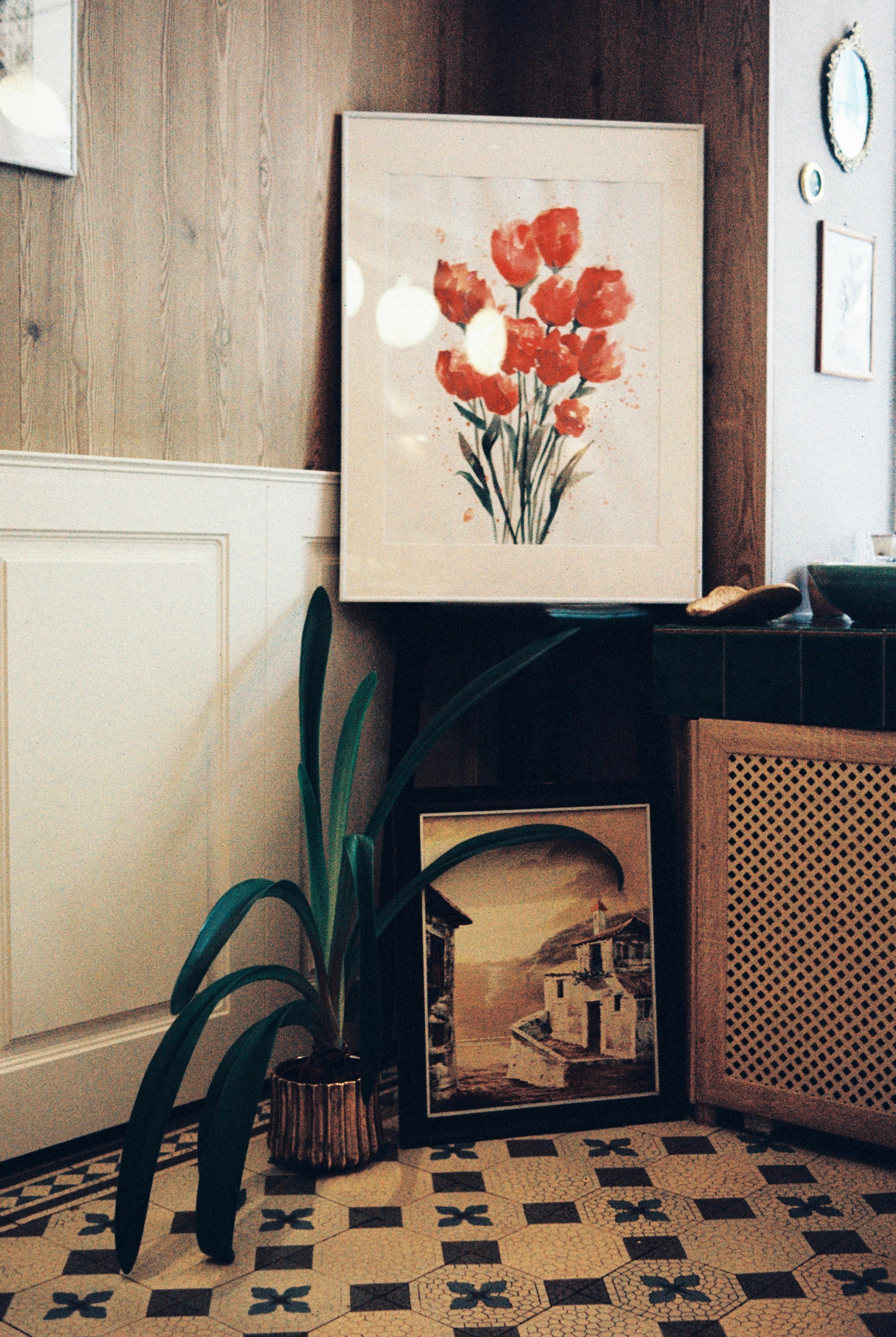 Framed red flower artwork sits above a plant-filled corner with decorative prints and vintage floor tiles in a softly lit interior.
