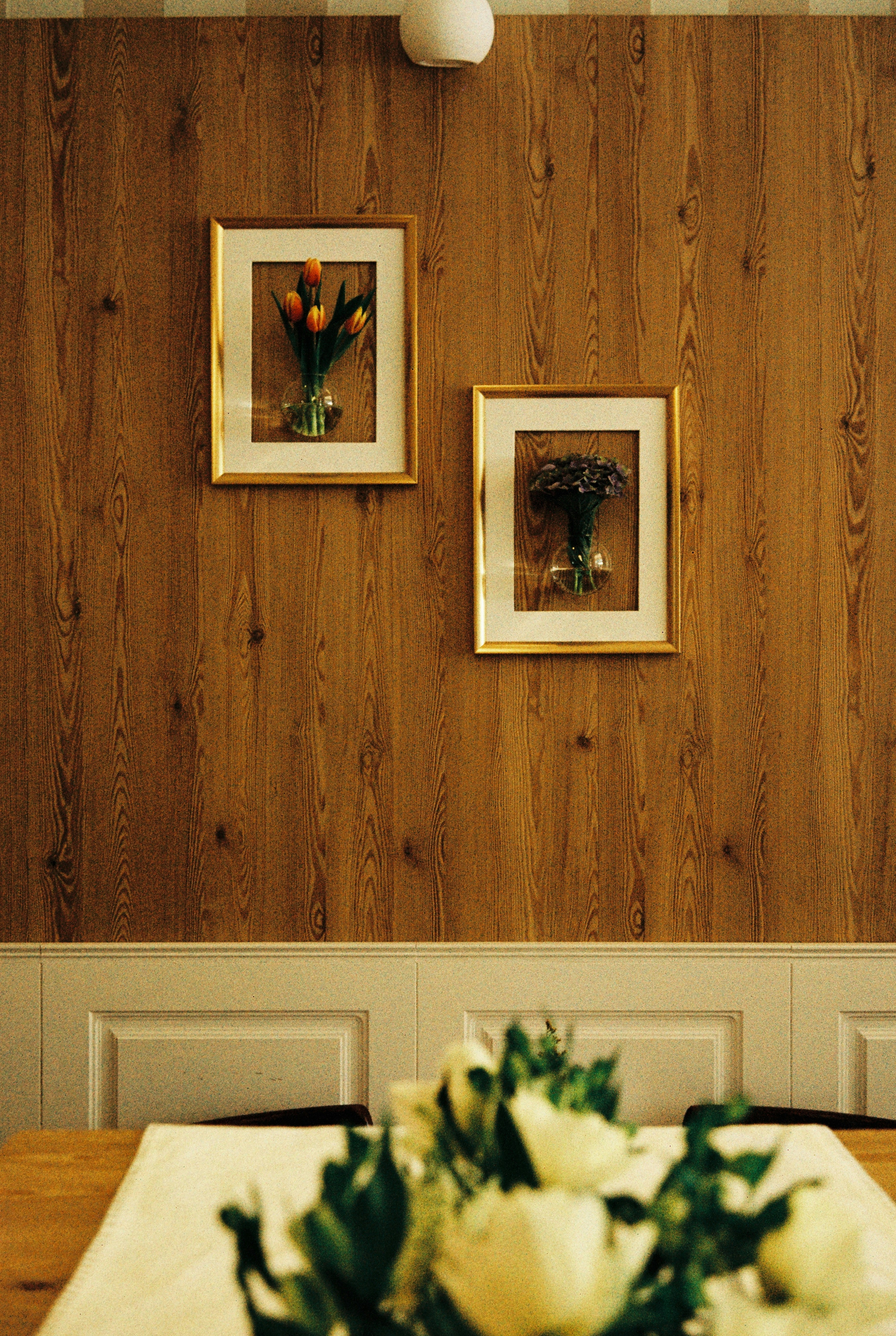 Wood-paneled wall with two framed floral photographs above a table setting featuring a white flower arrangement.