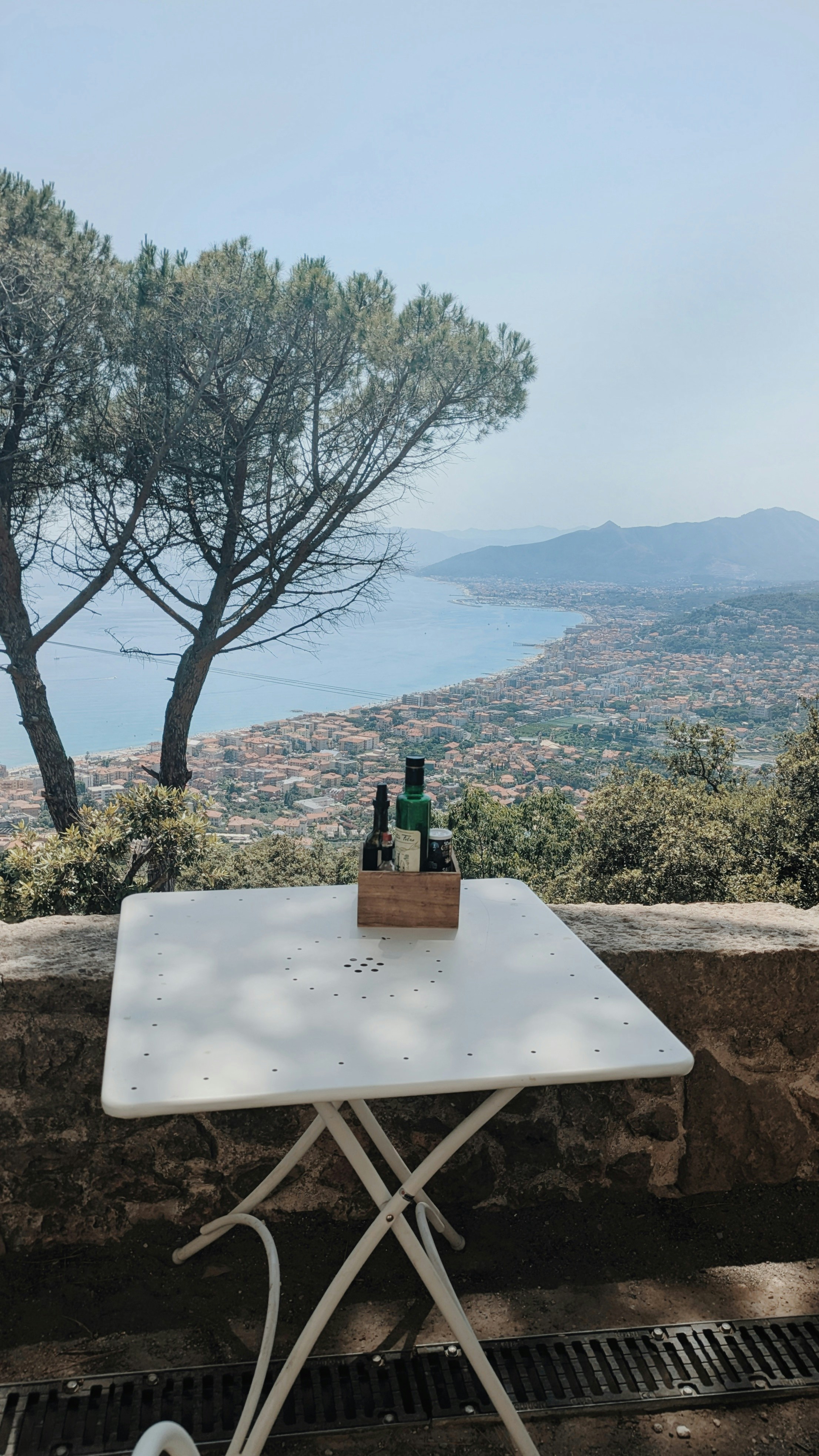 A white fold-out table with bottles sits on a stone ledge overlooking a coastal town and sea; the scene conveys a tranquil outdoor setting with distant hills and blue sky. This is a photograph, not a painting.