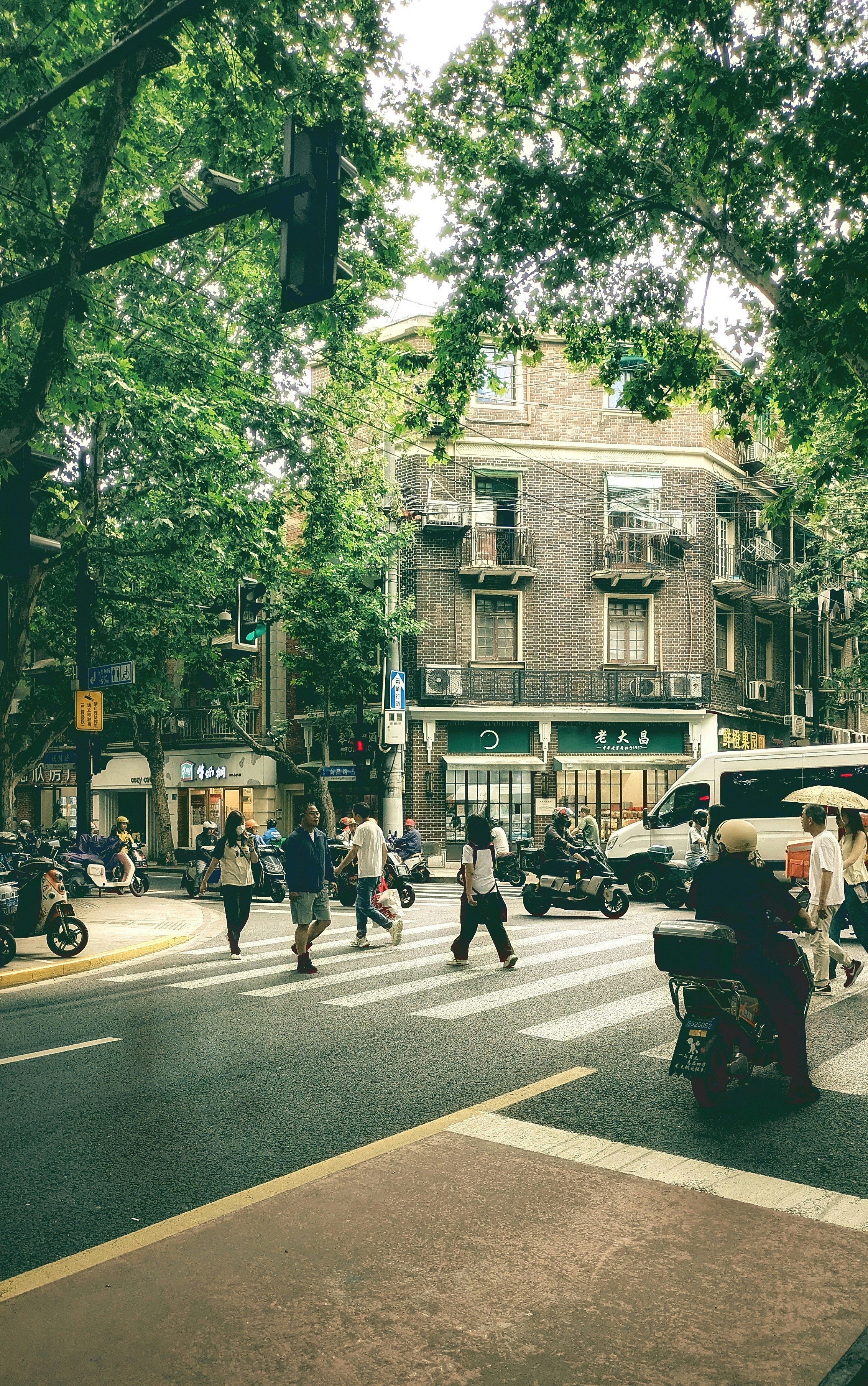 Bustling urban crosswalk beneath a canopy of trees; pedestrians and scooters glide by as a brick storefront forms the backdrop.